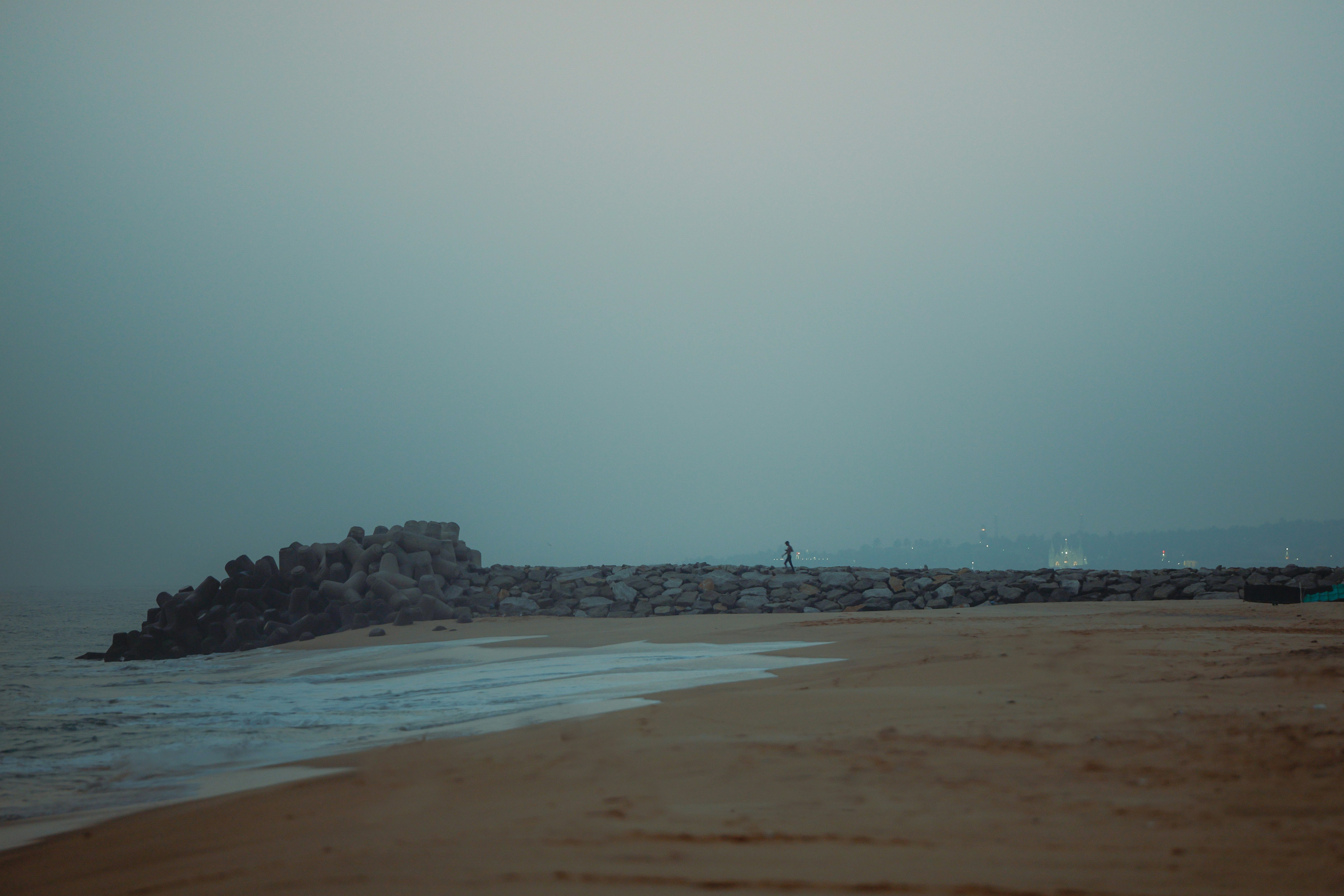 Lone figure walks along a rocky jetty under a misty sky, with soft waves lapping at a sandy beach.