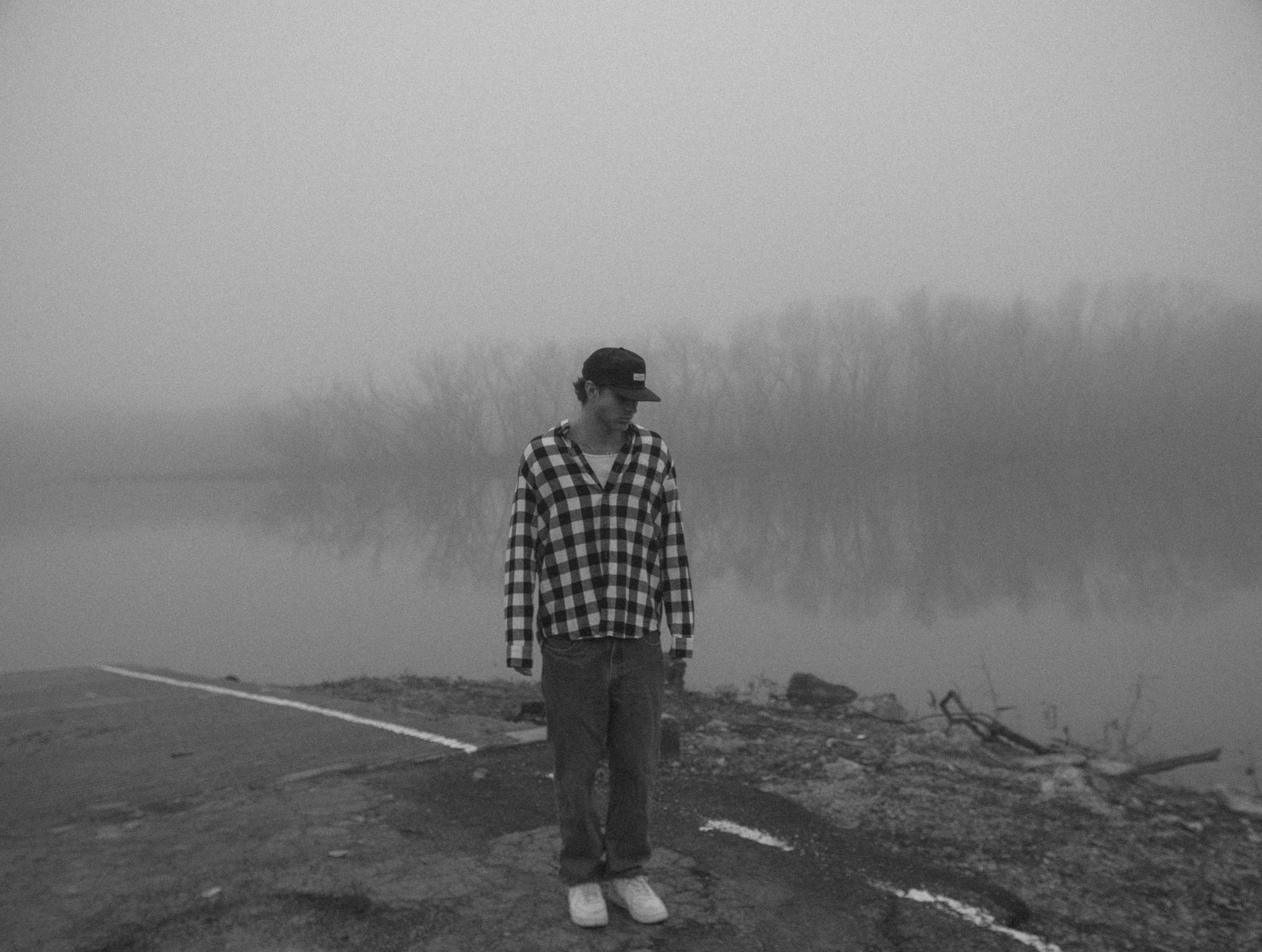 Solitary figure in a checkered shirt stands near a misty lakeside, surrounded by blurred trees and foggy ambiance.