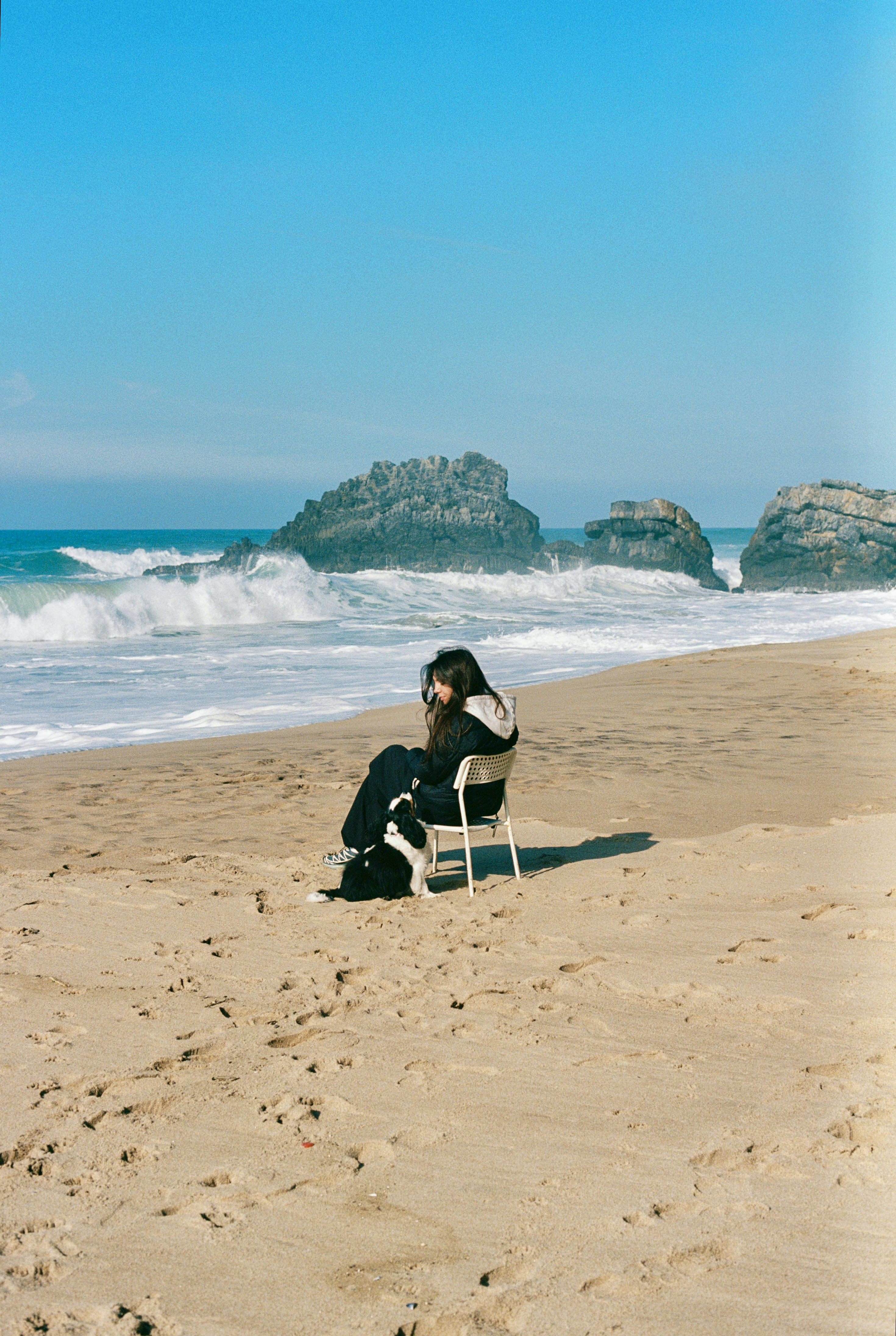 Candid photograph of a woman in dark clothing seated on a chair with a dog on a sunlit beach, as waves crash near rugged rock formations.