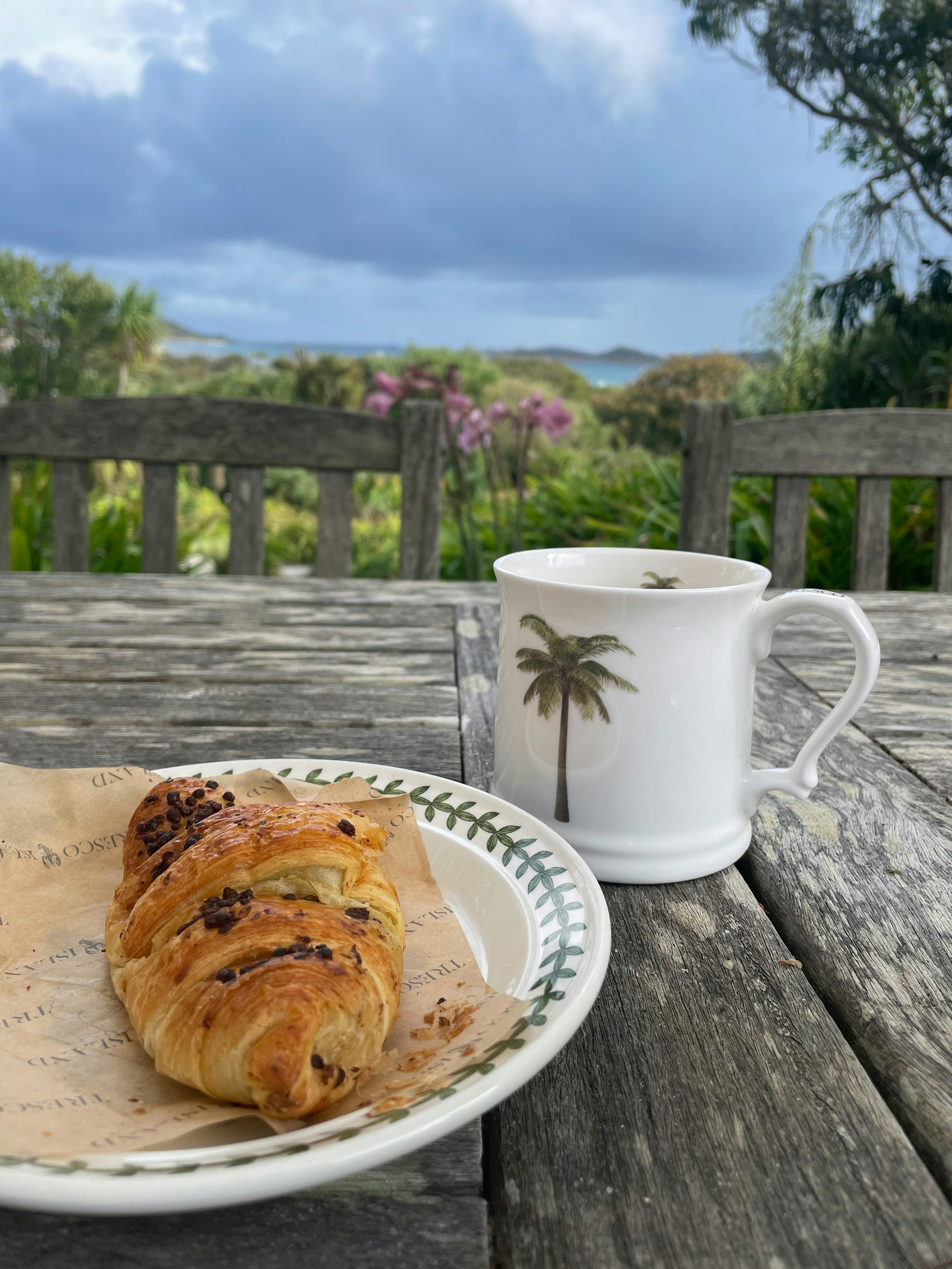 Rustic Coastal Breakfast: Croissant on Weathered Wood with Palm-Mug and Sea View