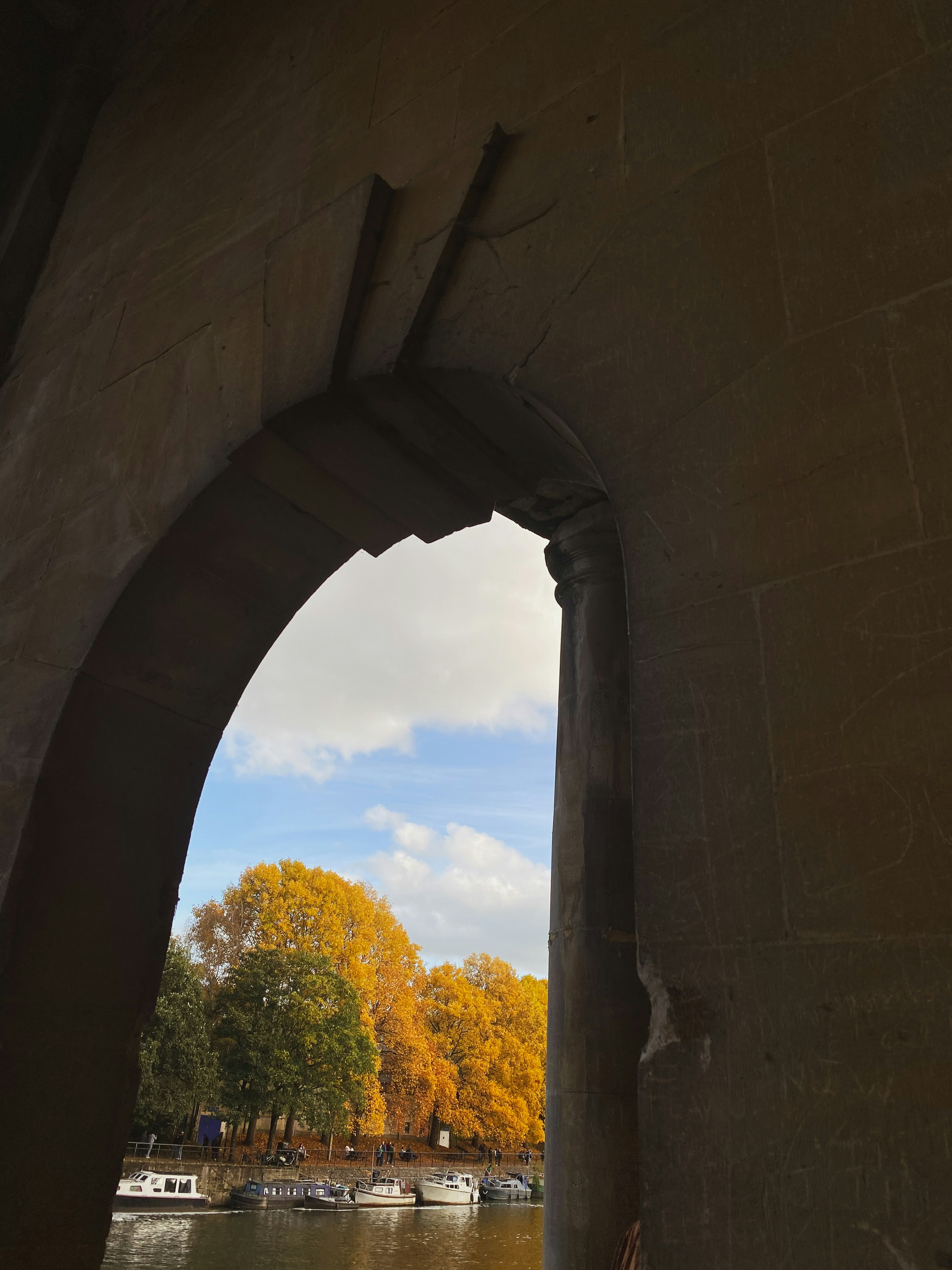 View through an archway showcasing vibrant autumn trees along a riverbank, with boats gently floating on the water.