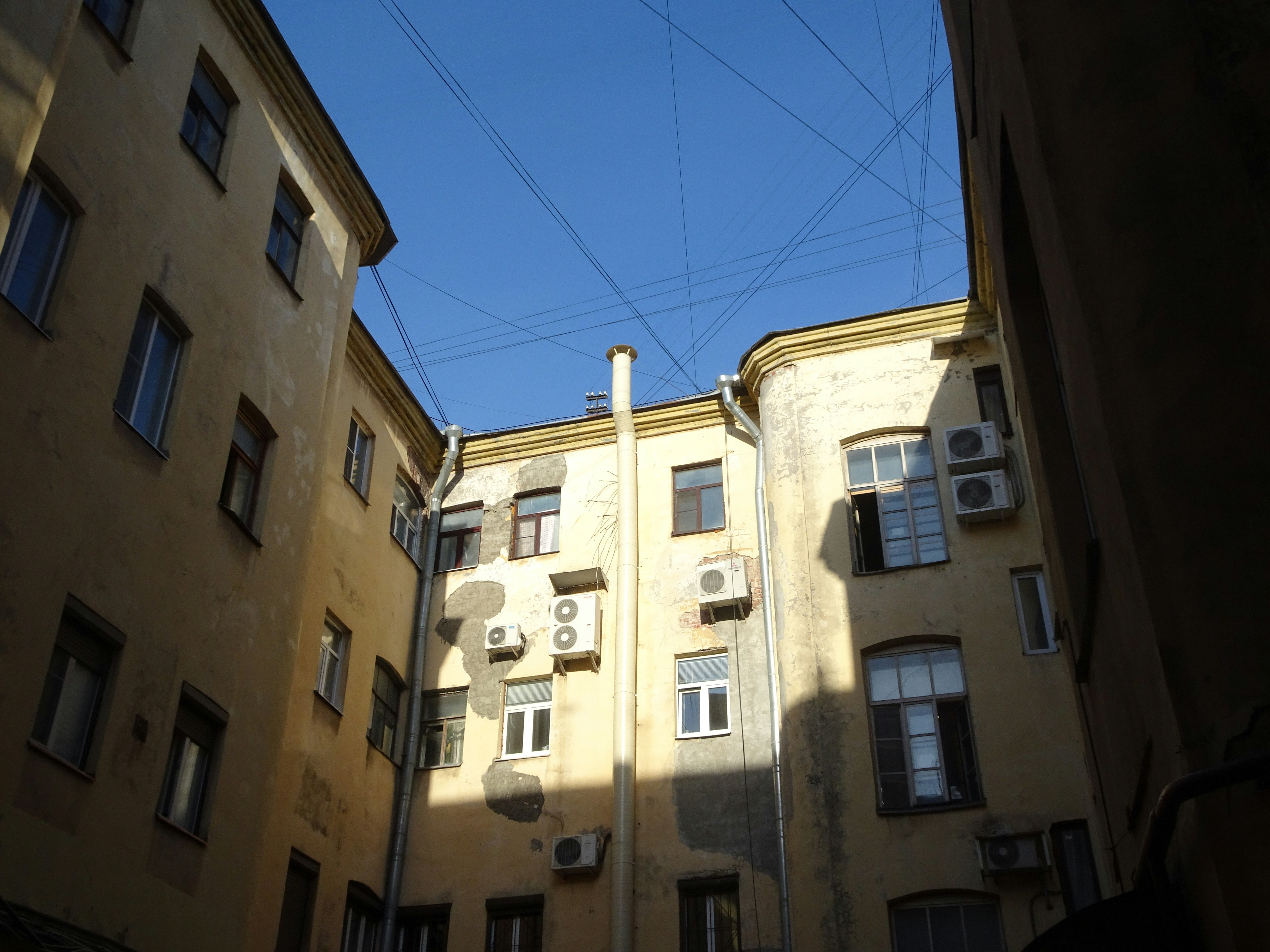 Sunlit central wall of a weathered courtyard building, with rows of windows and visible air-conditioning units against a clear blue sky.