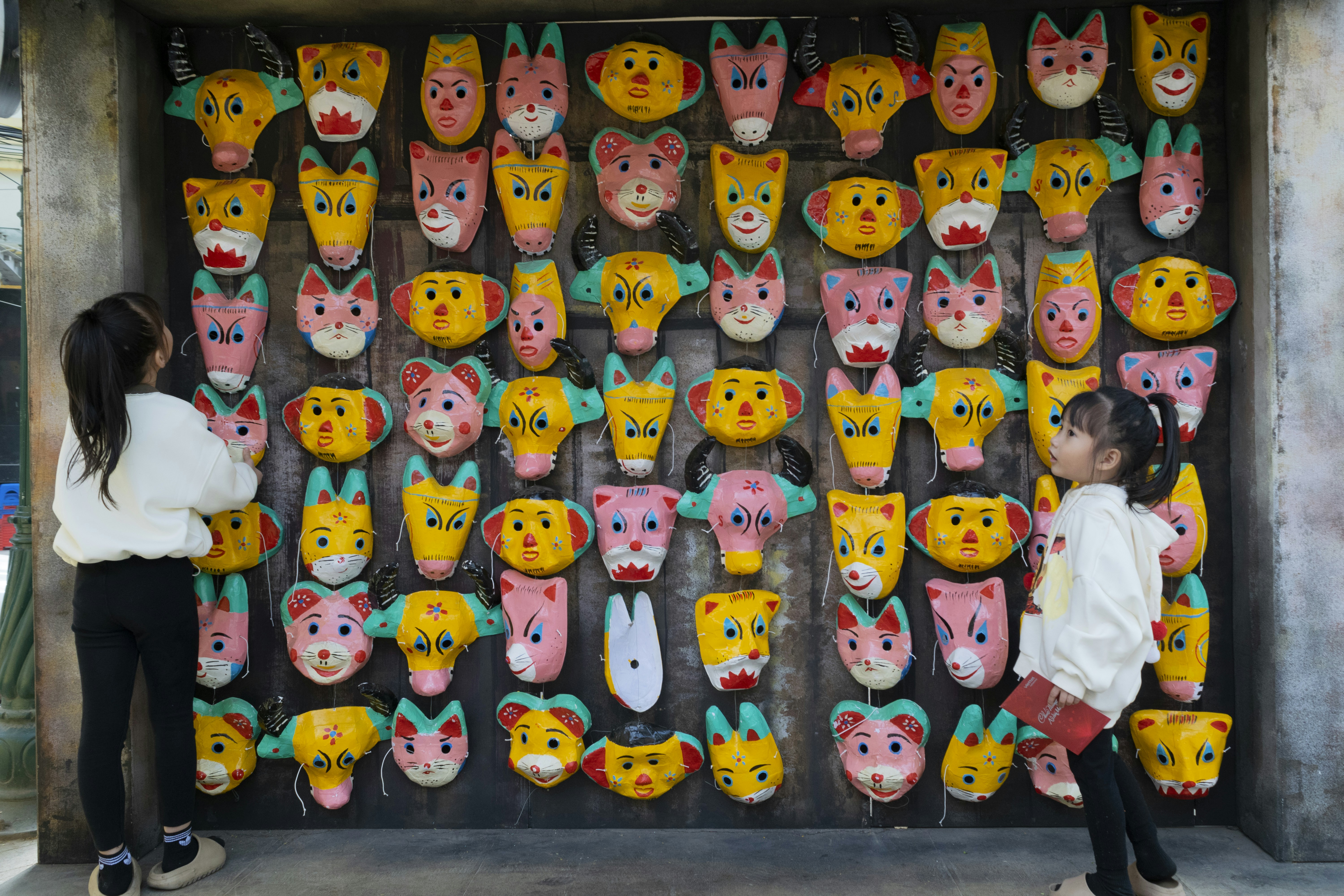 Children explore a wall of colorful masks painted in vivid yellows, pinks, and greens, creating a lively scene.