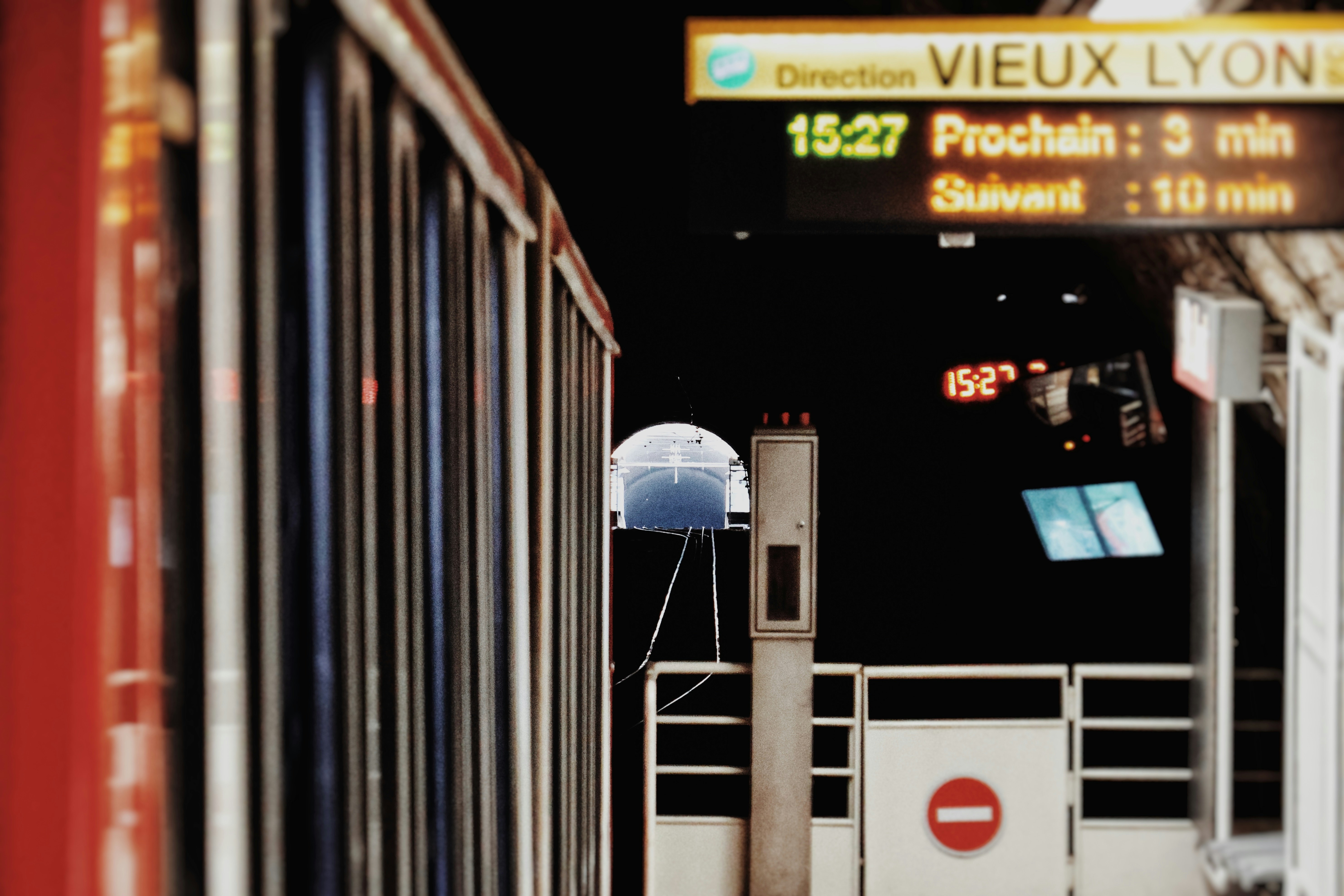 Train station platform with digital display showing upcoming train times and a train partially visible on the left.