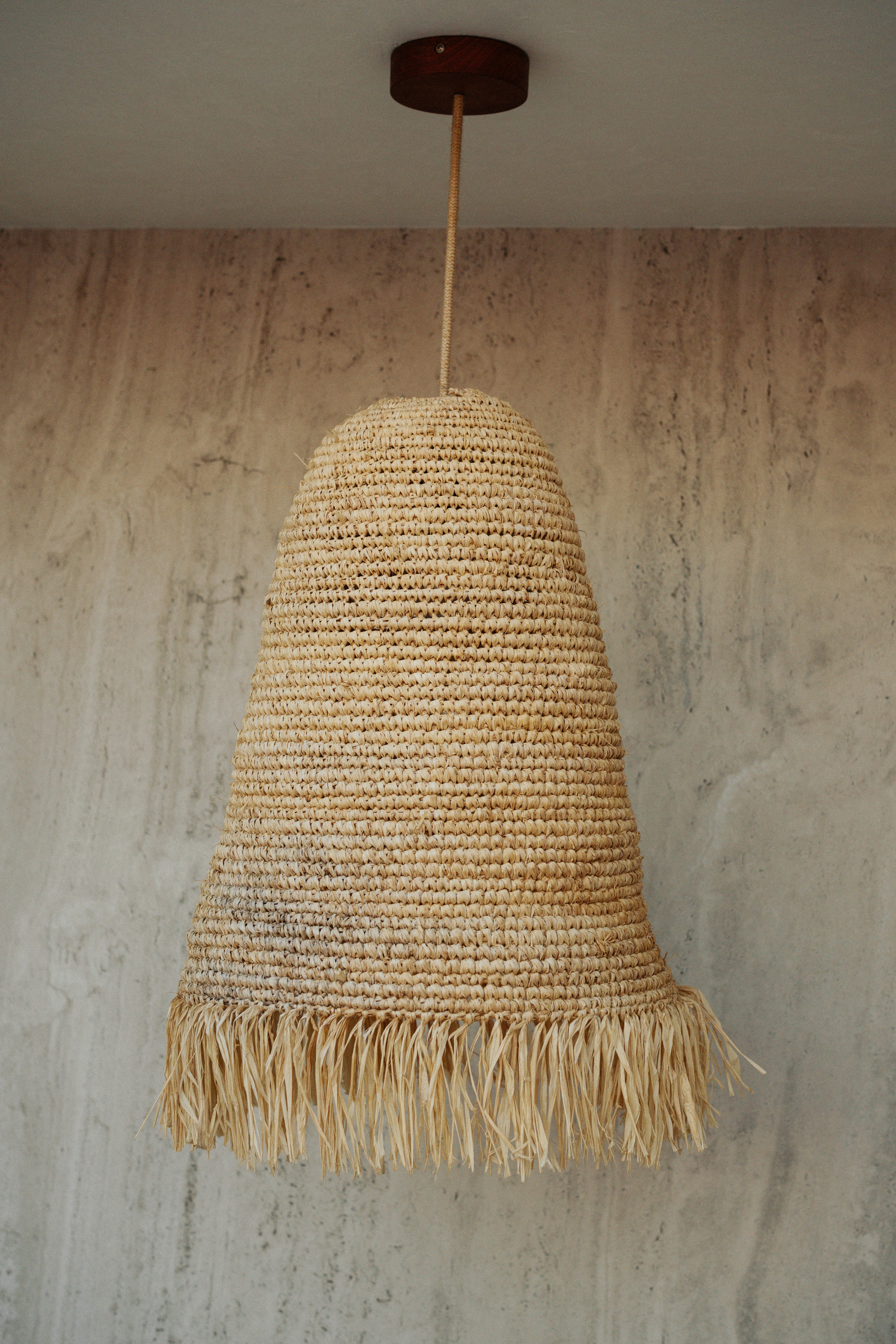 A handwoven straw pendant light hangs from the ceiling against a textured concrete wall. The warm, diffuse glow highlights the natural fibers and fringe, emphasizing artisanal detail.
