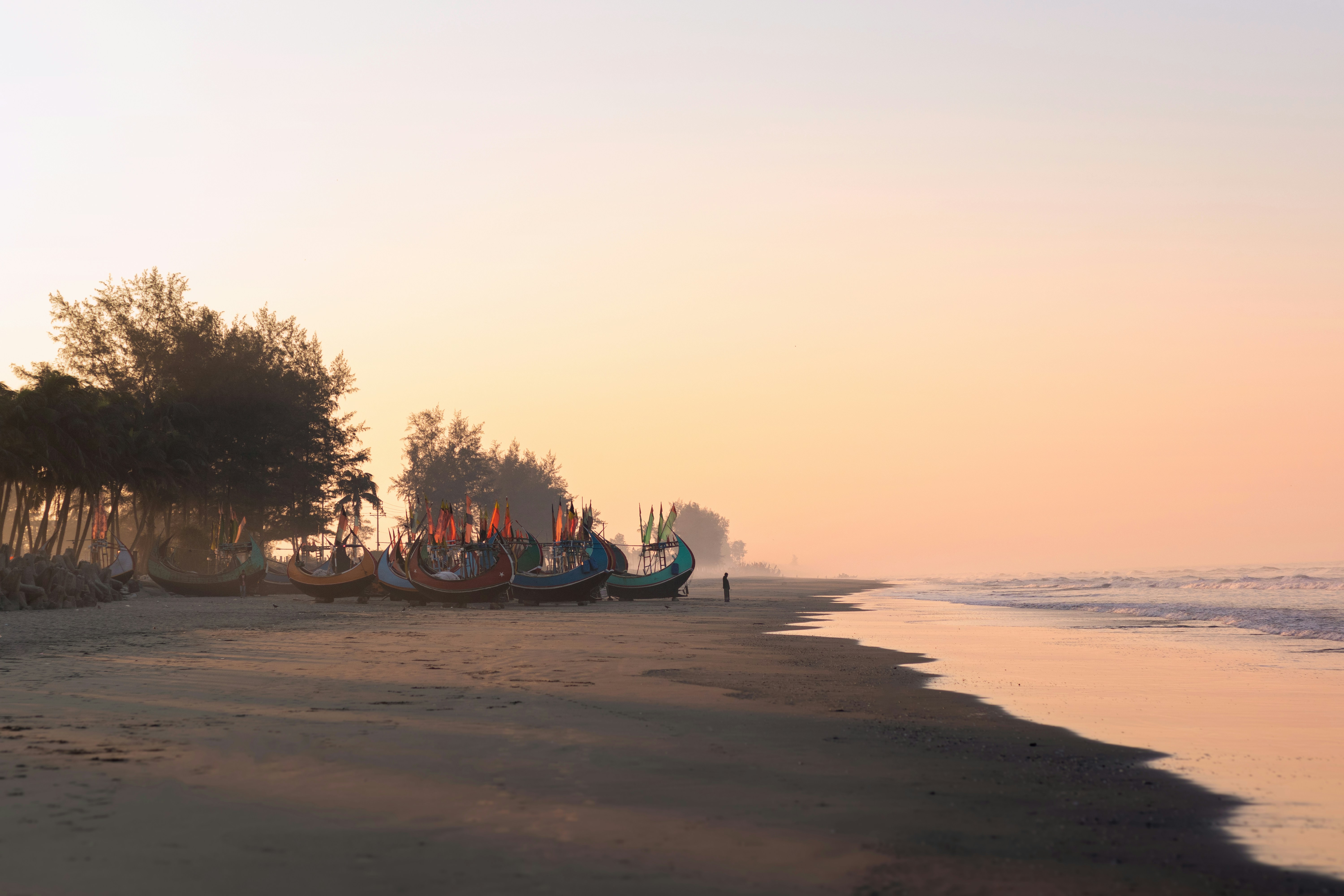 Colorful fishing boats rest on a sandy beach at sunrise, framed by silhouetted trees and pastel skies.