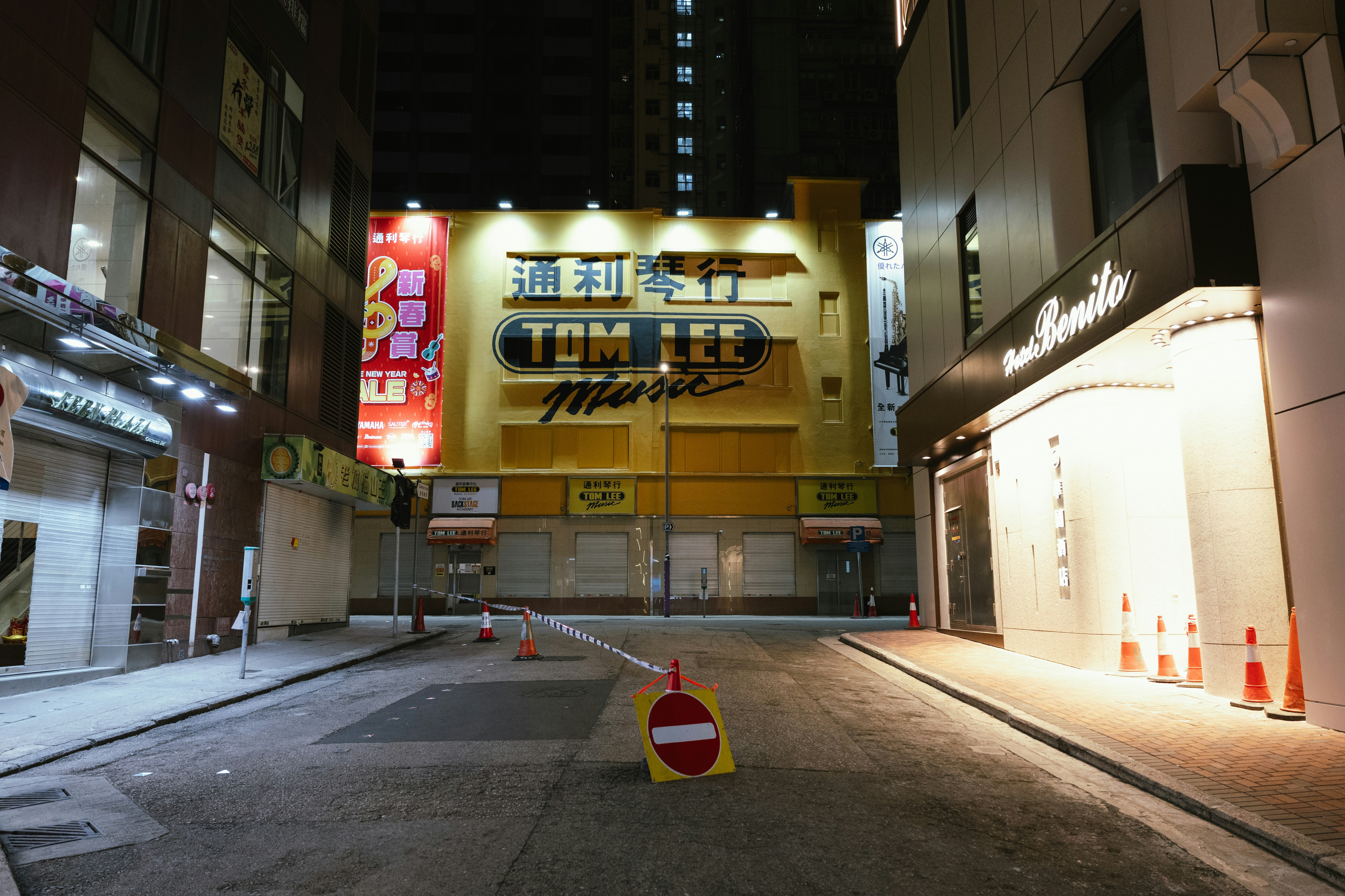 Deserted urban alley at night with a vibrant yellow music store facade, surrounded by muted buildings and illuminated by artificial lights.