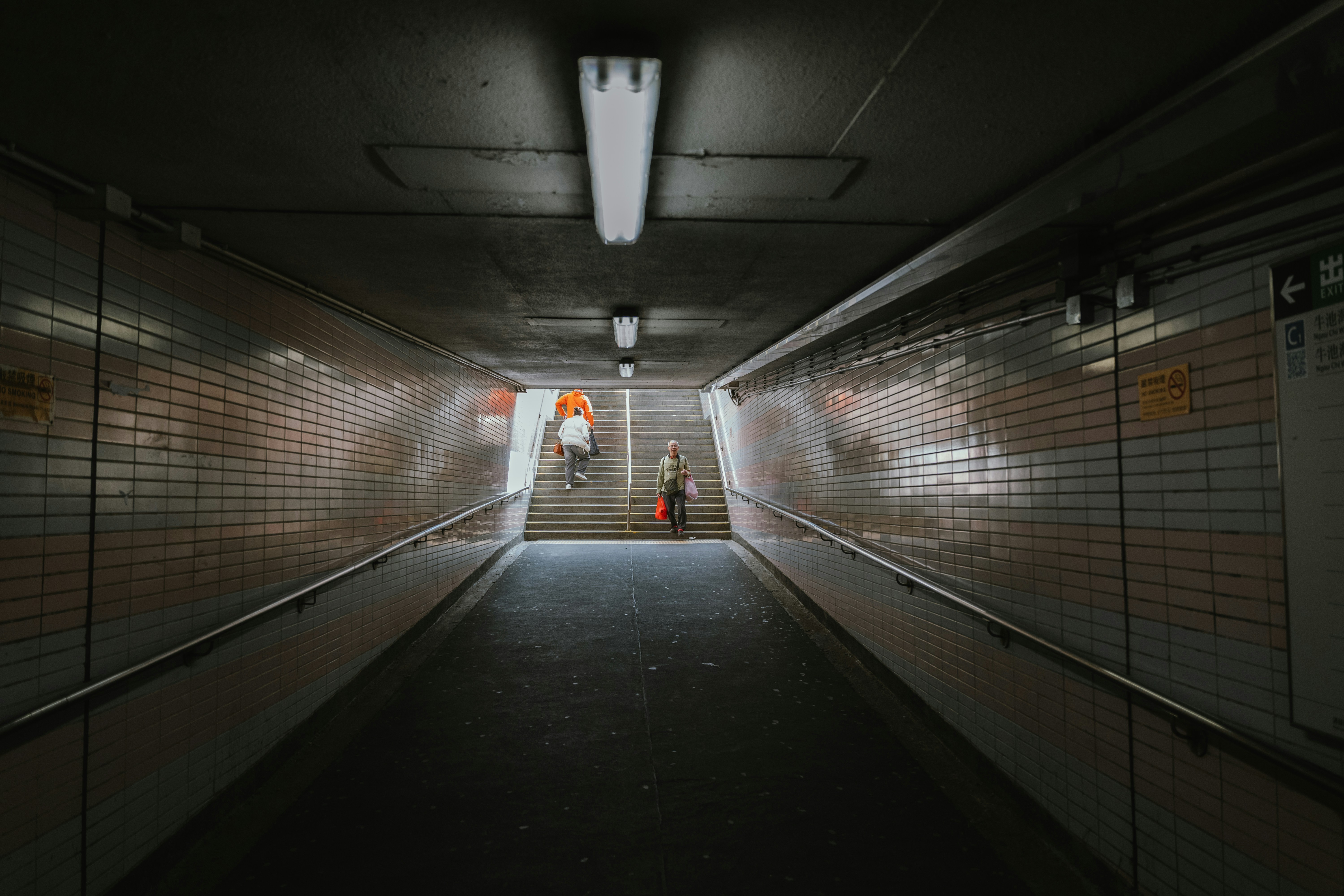 Dimly lit underground passageway with two figures ascending stairs, one in vibrant orange contrasting the muted tiled walls.