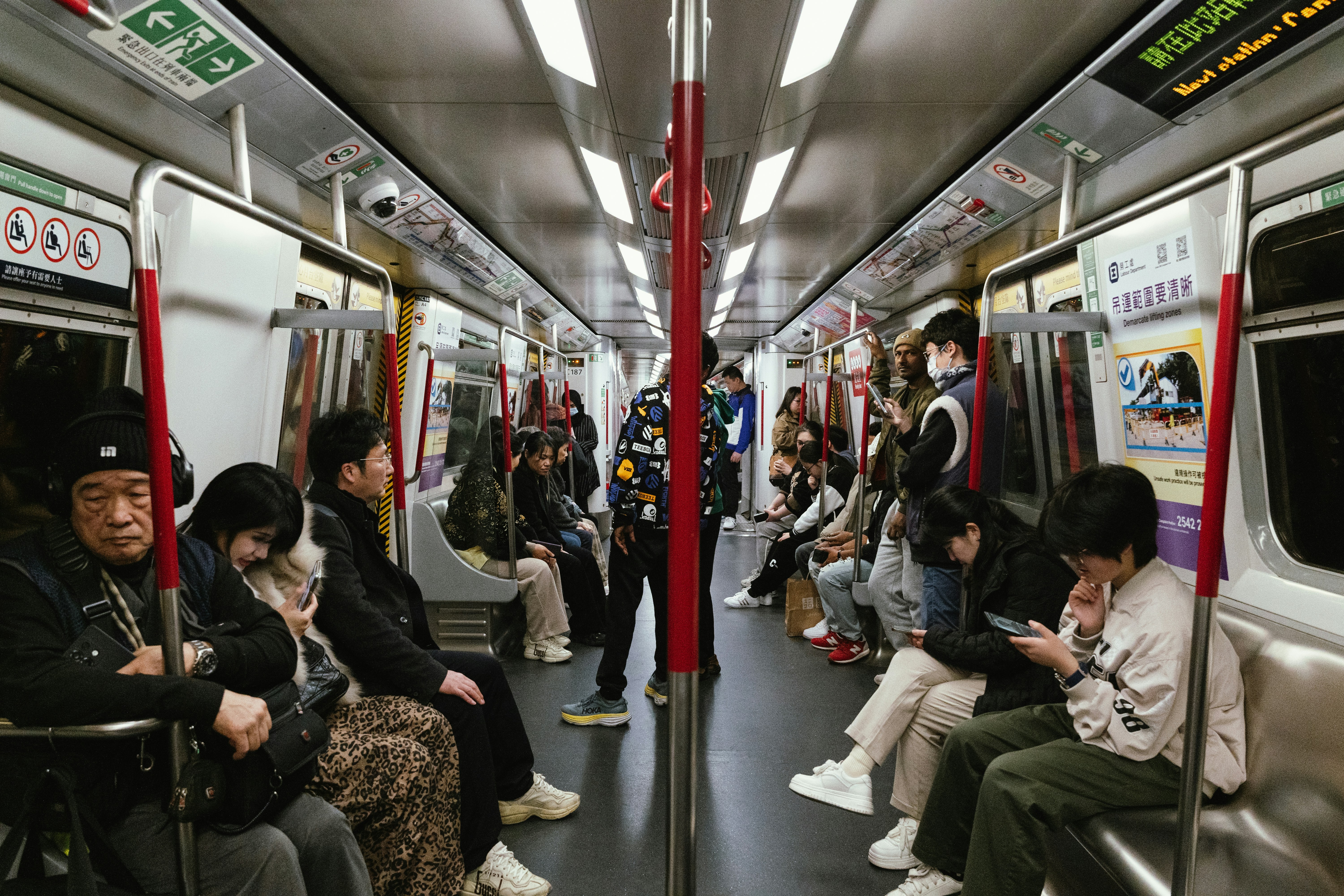 Commuters seated and standing inside a subway car, framed by red poles and overhead lighting.