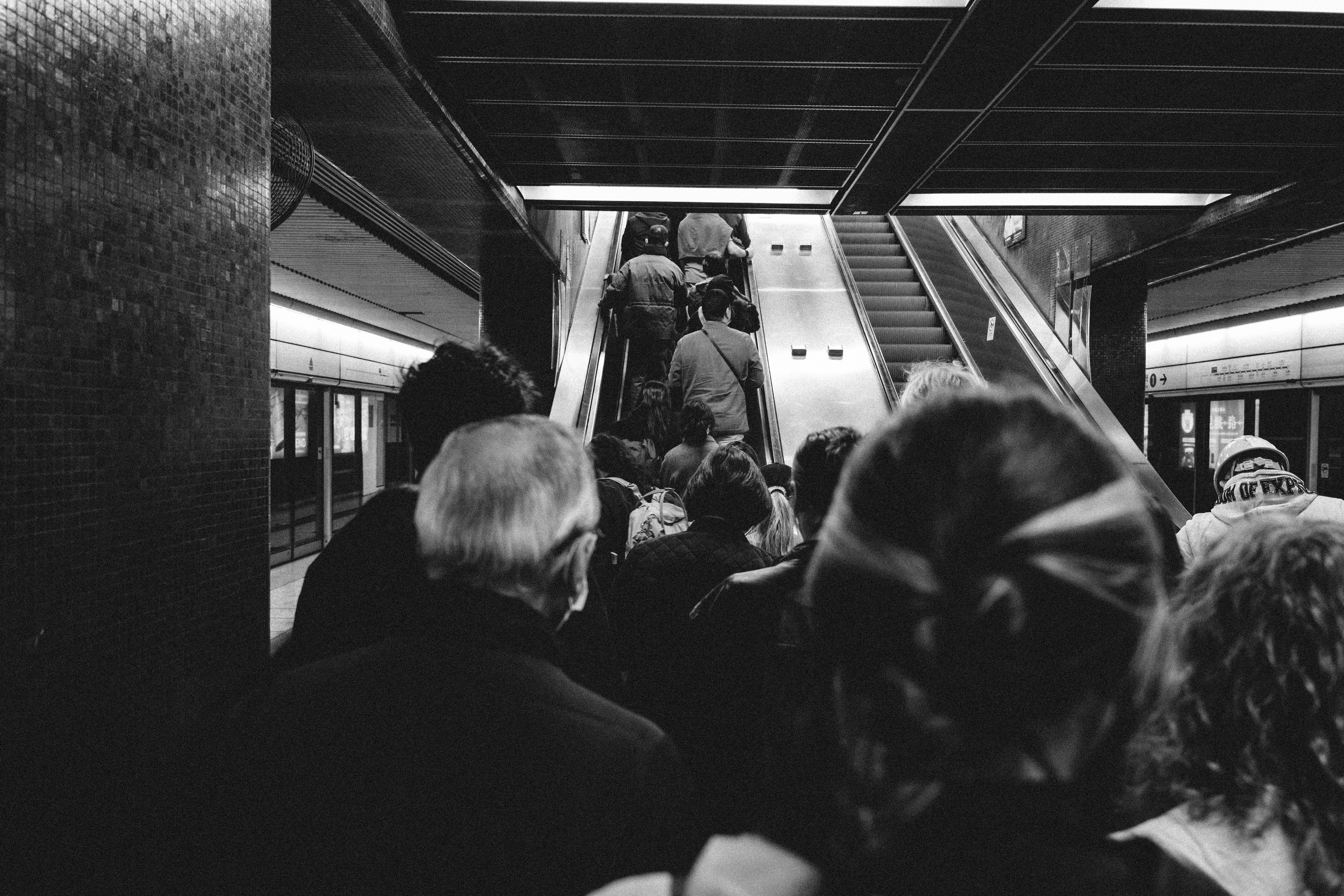 Crowd moving up an escalator in a metro station, framed by a strong overhead structure with contrasting silhouettes and bright lighting.