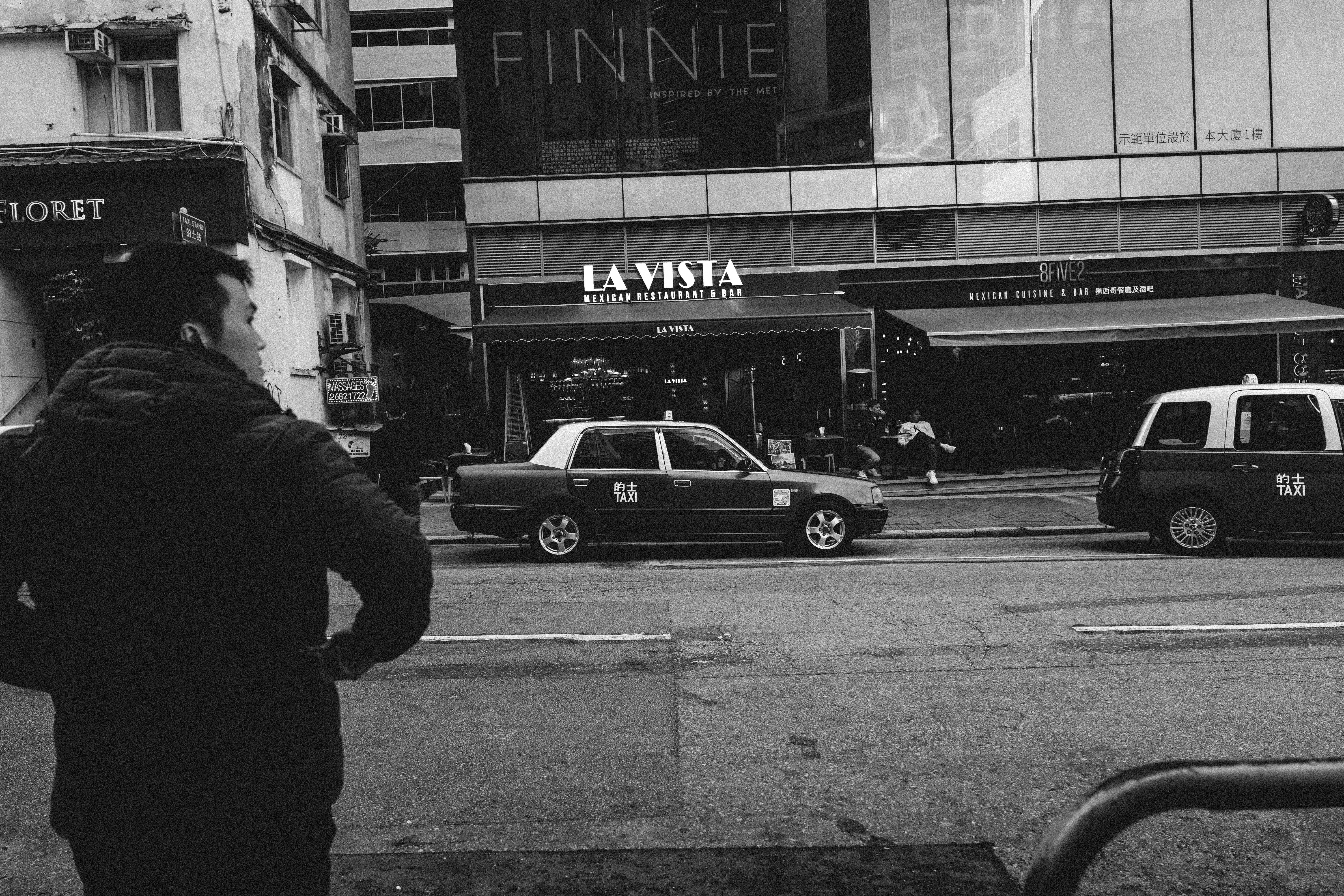 Monochrome urban street scene with a person observing taxis lined up across storefronts, balanced by building and road lines.