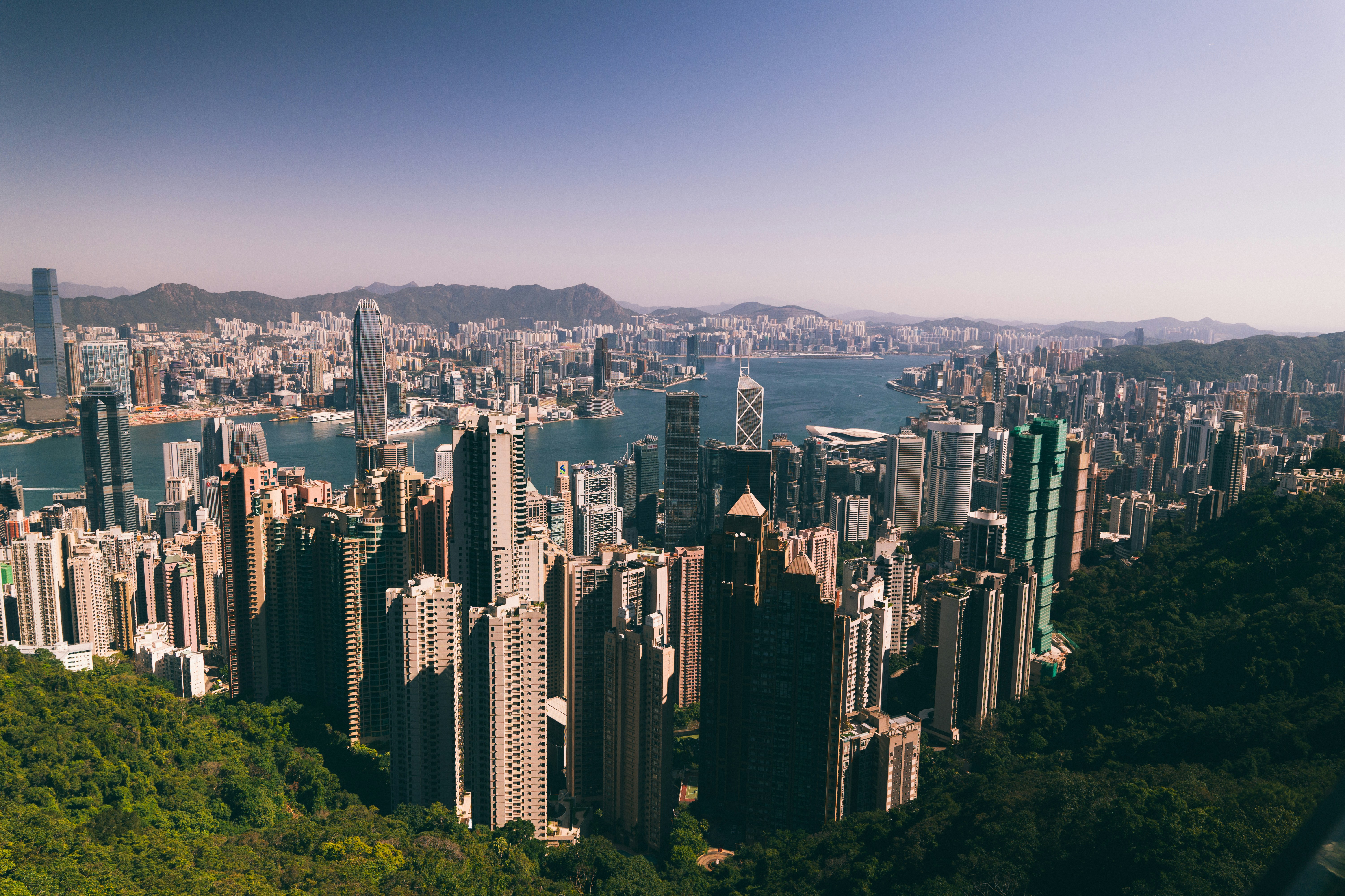Hong Kong skyline with tall skyscrapers and lush hills under a clear sky.