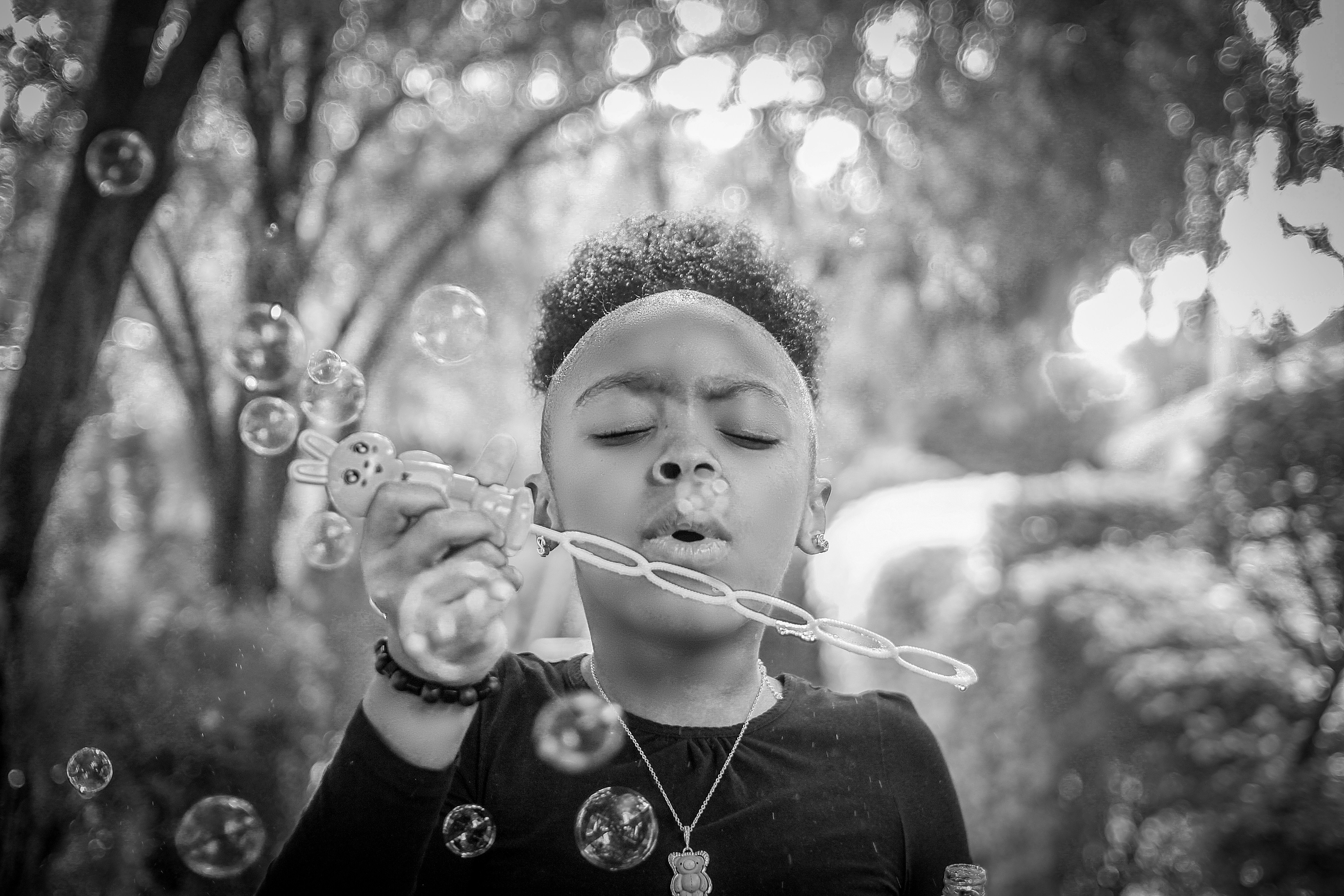 Child blowing bubbles with closed eyes, surrounded by soft bokeh from sunlight through trees.