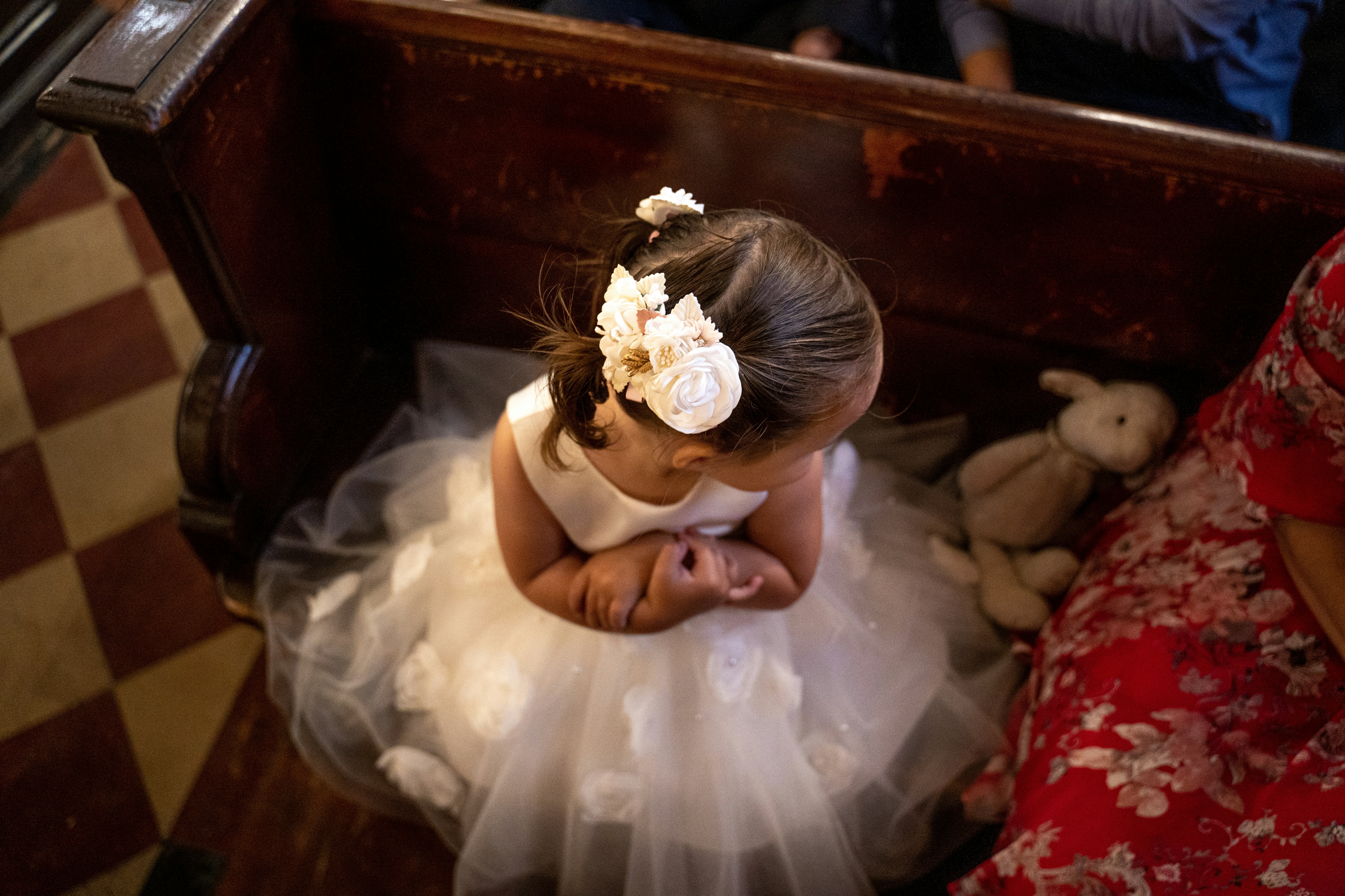 Child in white tulle dress with floral hairpiece seated in a wooden pew beside a plush toy.