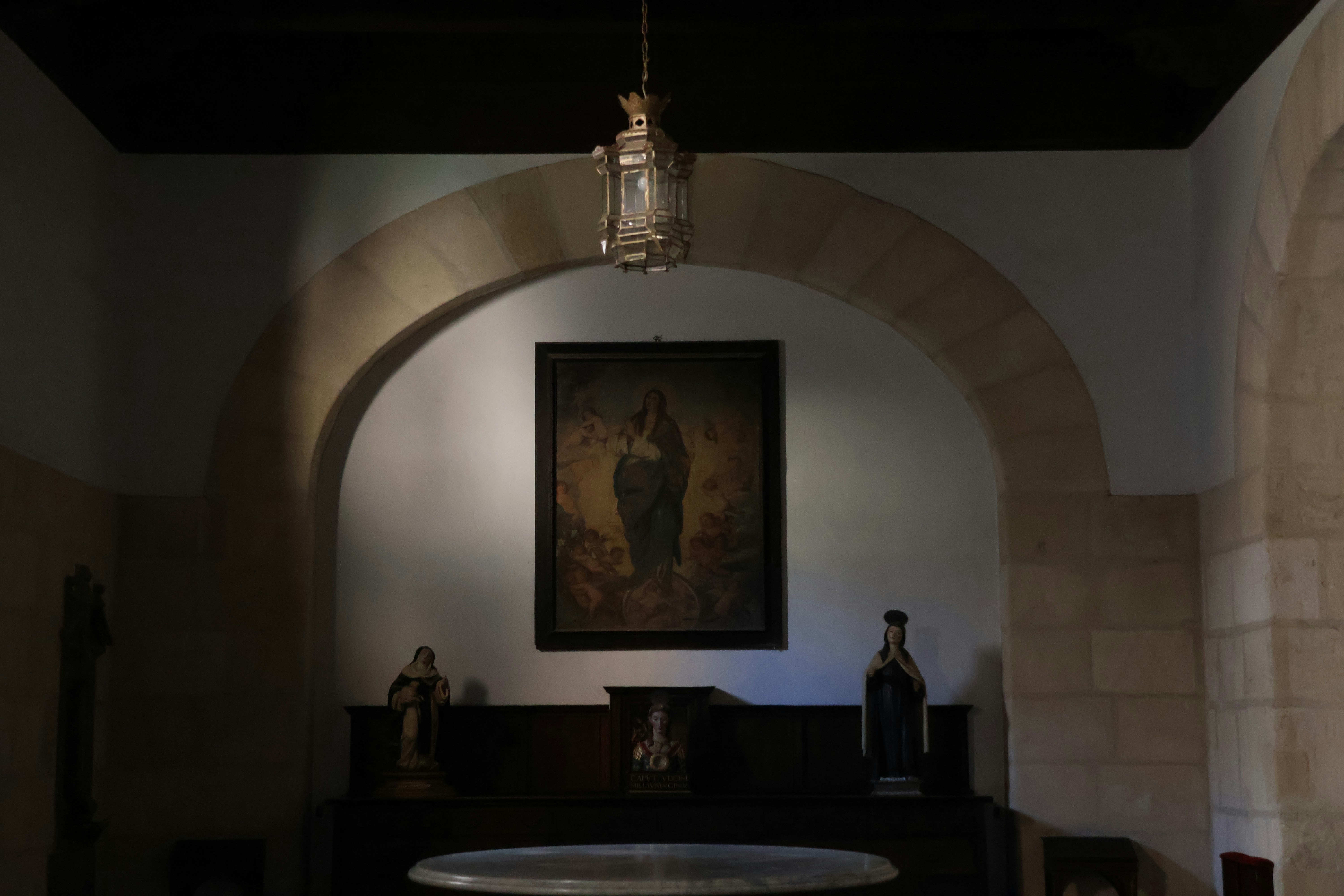 Dimly lit chapel interior with a central painting, flanked by religious statues under a stone arch.