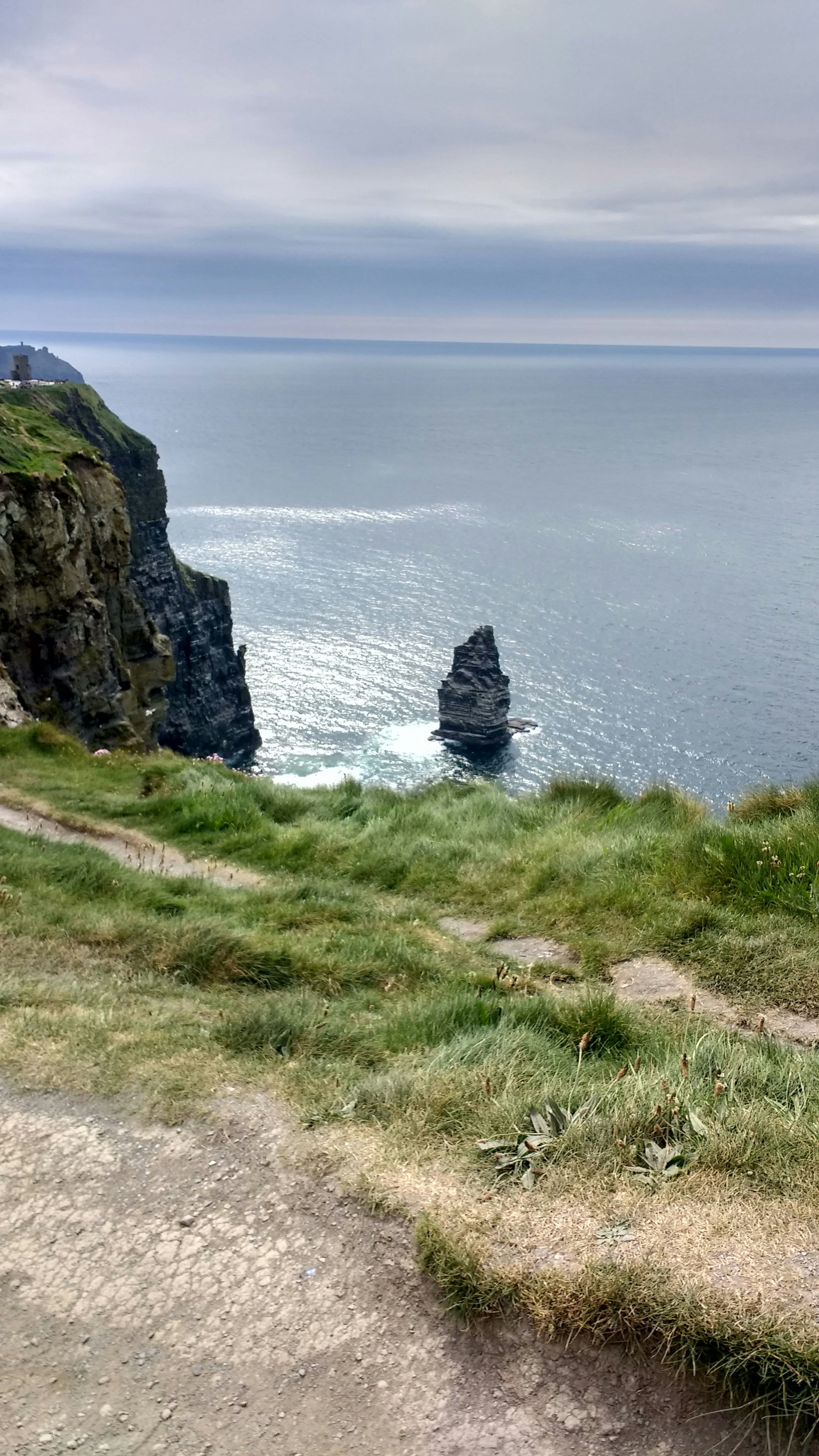 A rugged coastal cliff overlooking a solitary rock formation rising from the ocean. The gentle waves reflect the cloudy sky above.