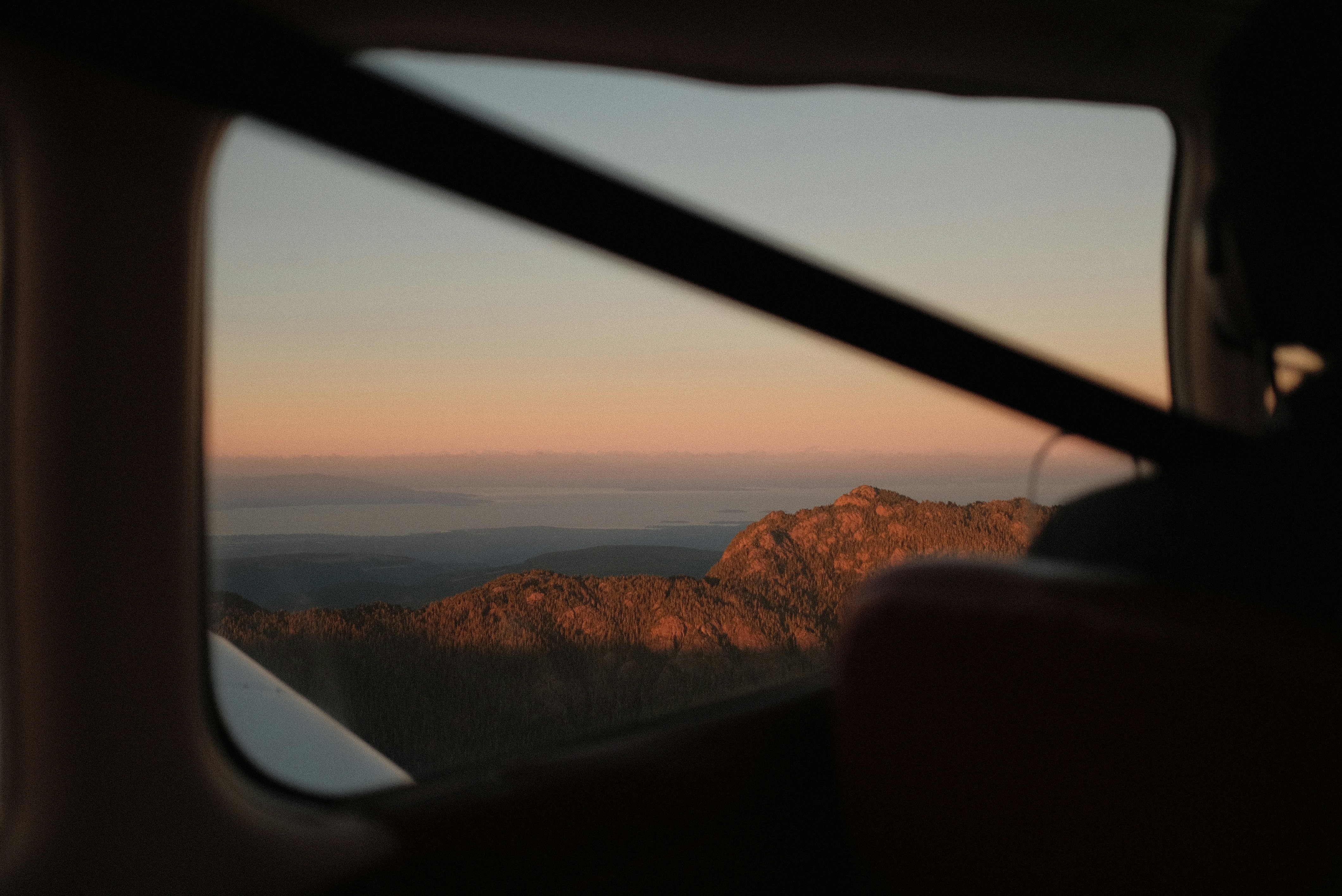 Mountains bathed in sunset hues viewed through a plane window, with a silhouetted seatbelt adding depth.