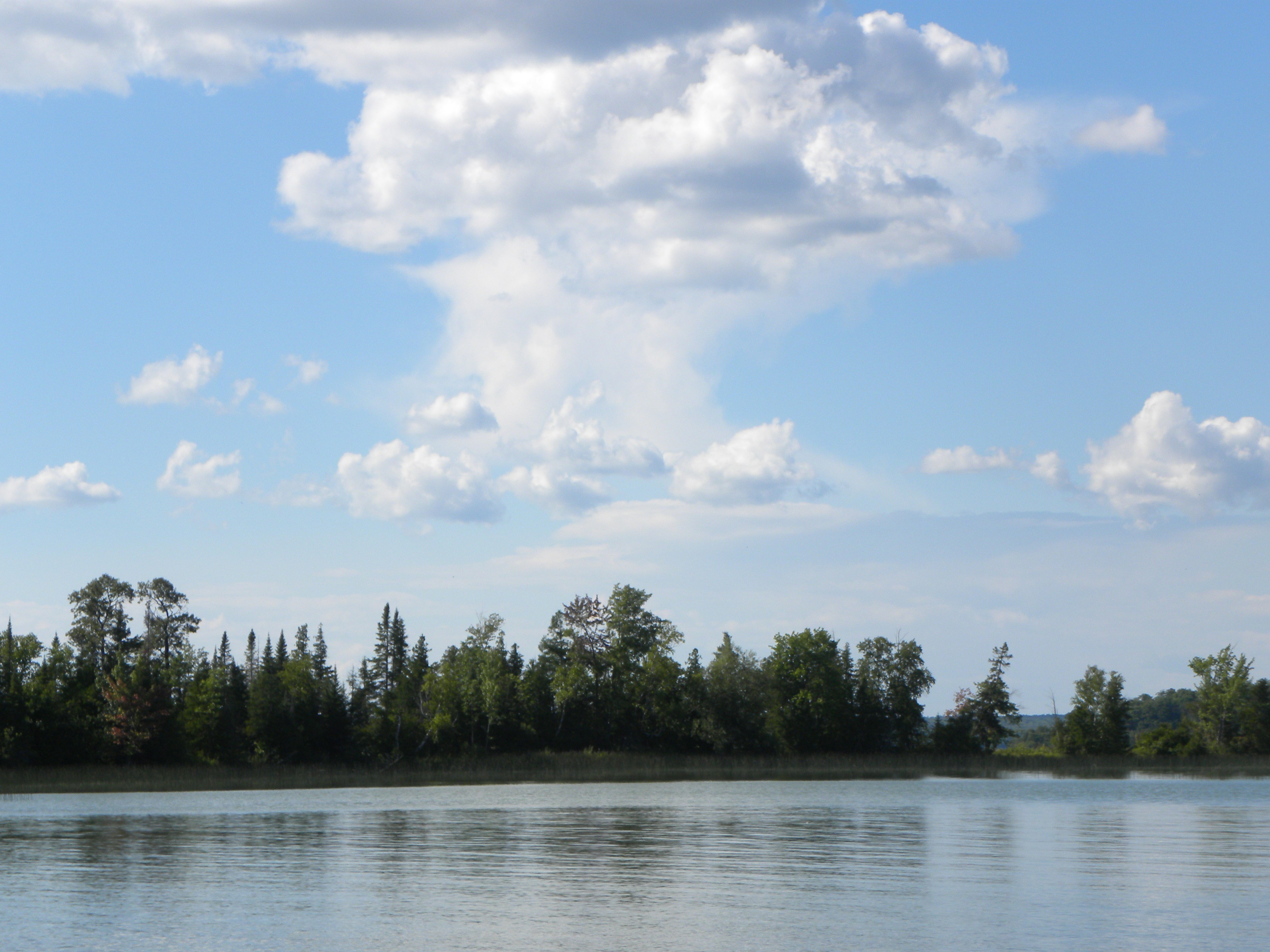 Dramatic cloud formation rises above a tranquil lakeside with lush green trees and calm reflective waters.