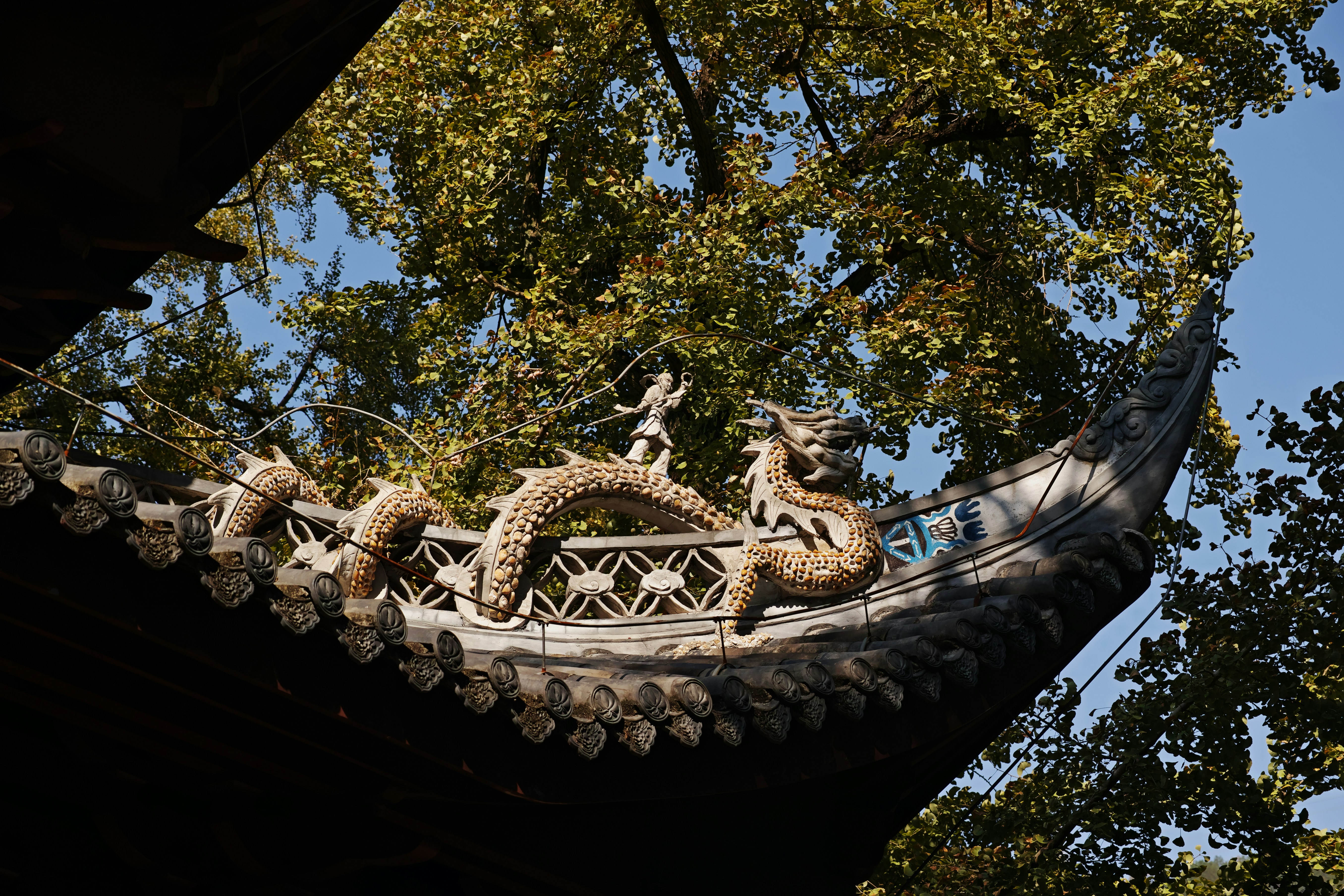 Dragon ornament along a traditional temple roof edge, photographed against a backdrop of green leaves and a clear blue sky.
