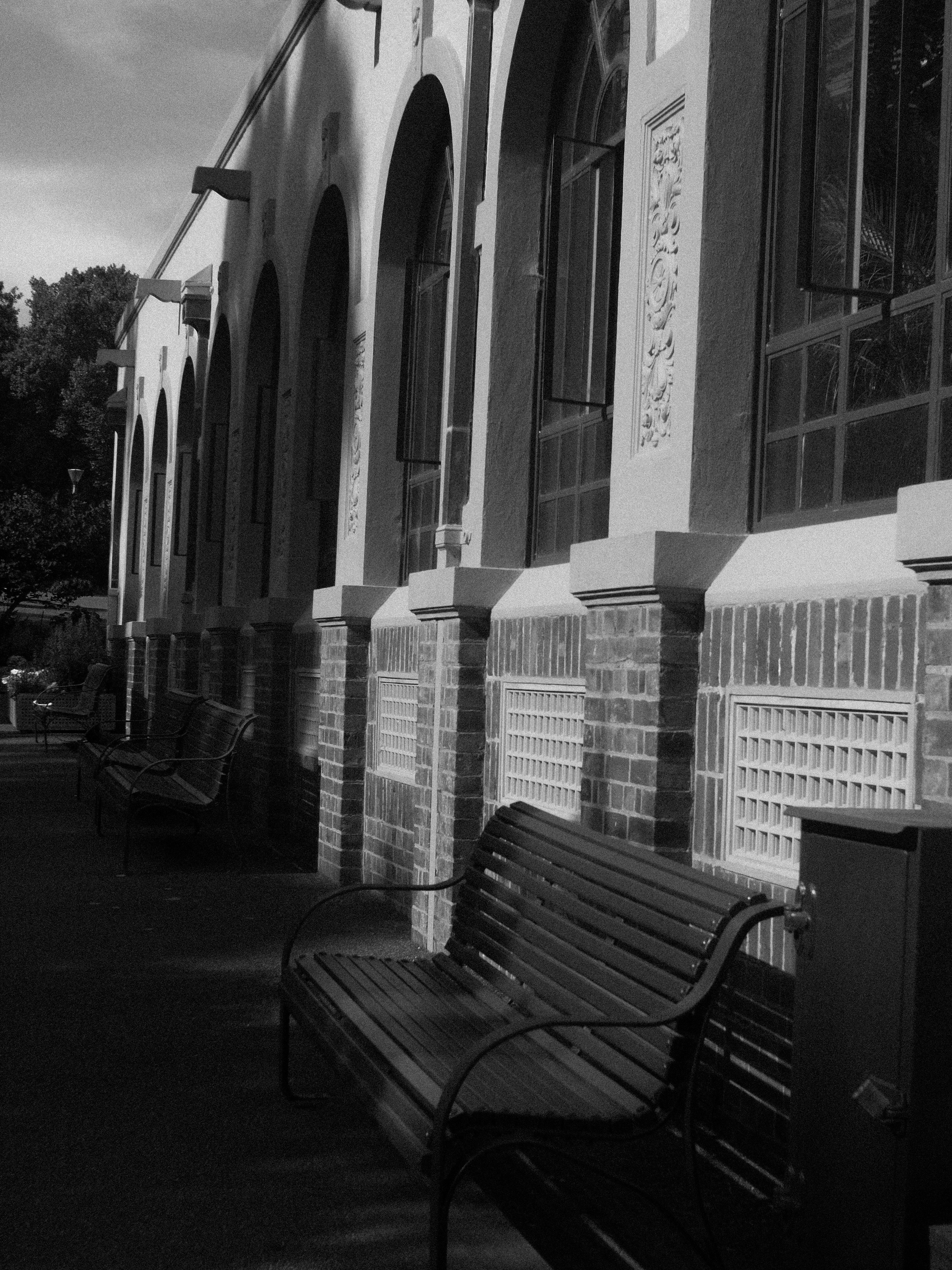 Black and white photo of a long row of arched windows and benches along a brick building facade.