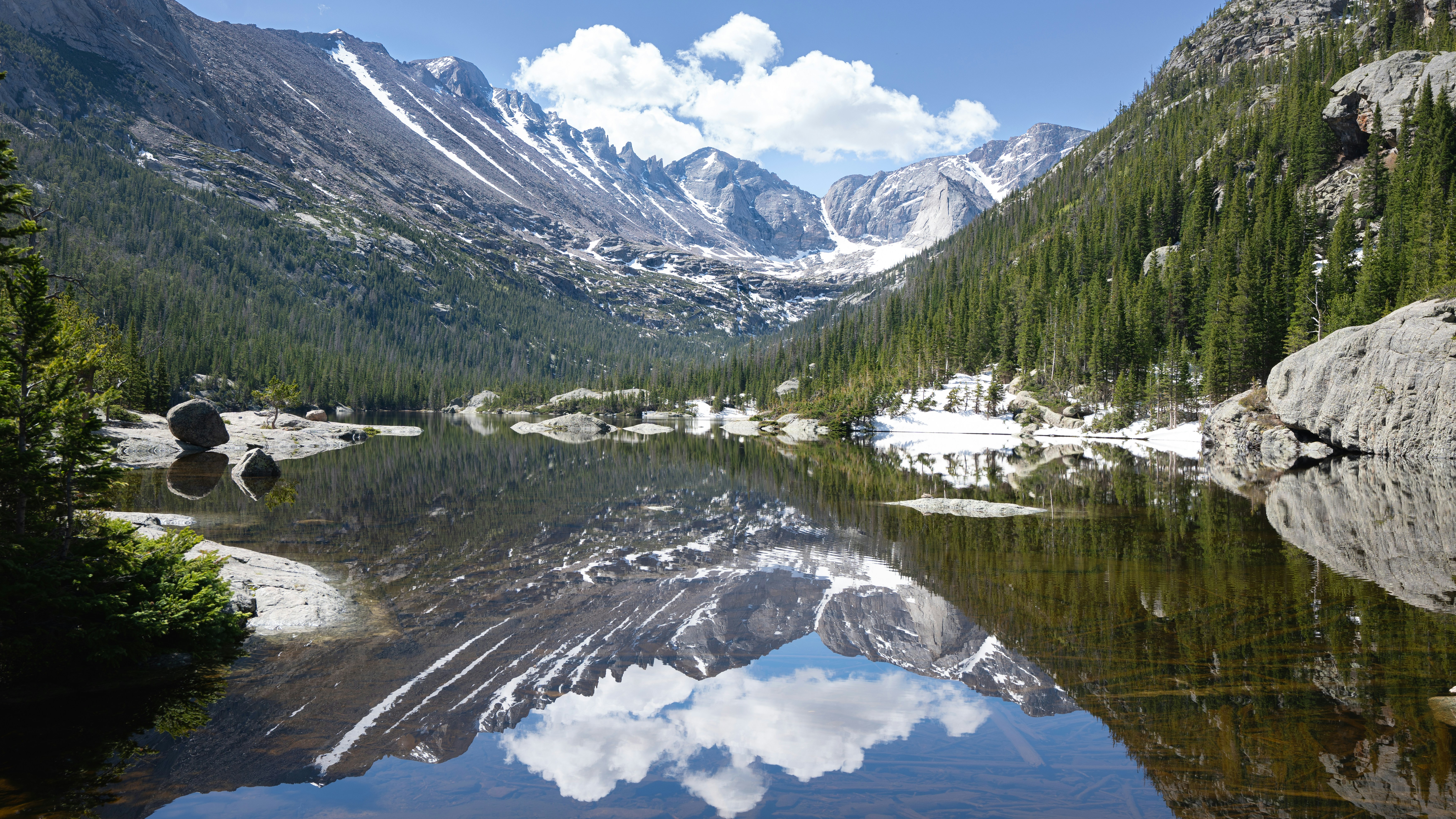 Alpine lake reflecting snow-capped mountains and pine forests under a bright sky with clouds.