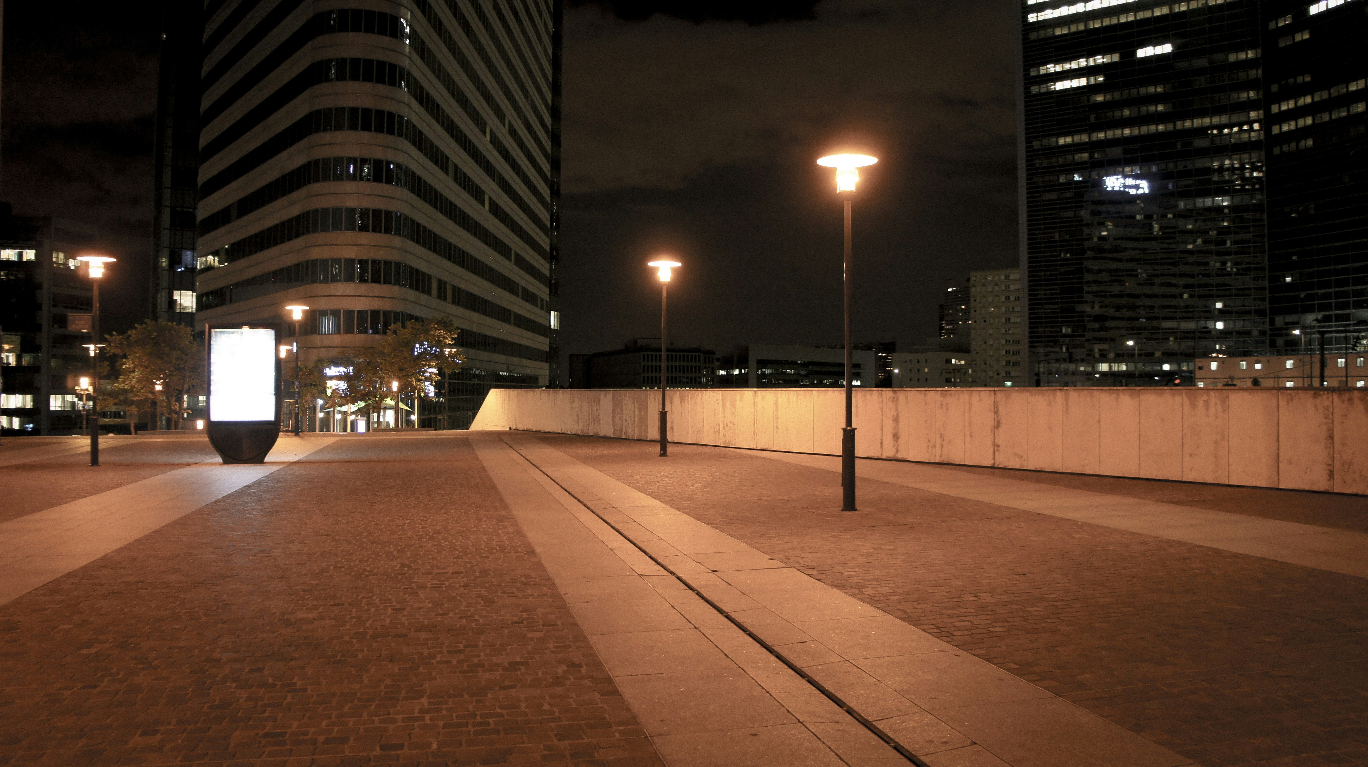 Urban walkway lit by street lamps, casting warm light on the brick pavement with modern buildings silhouetted against a dark sky.