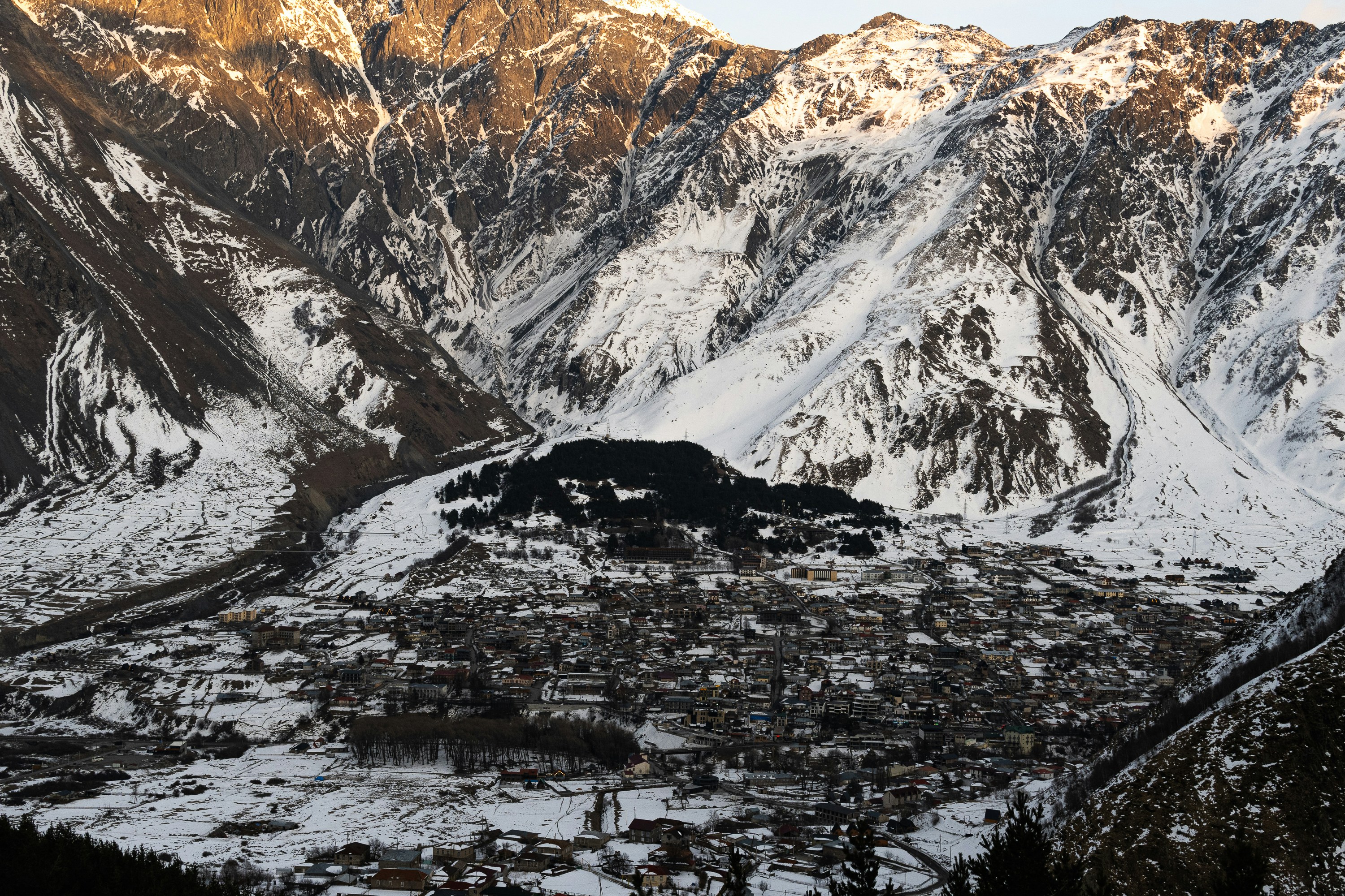 Mountain village nestled beneath snow-covered peaks with golden light on the rugged slopes at dusk.