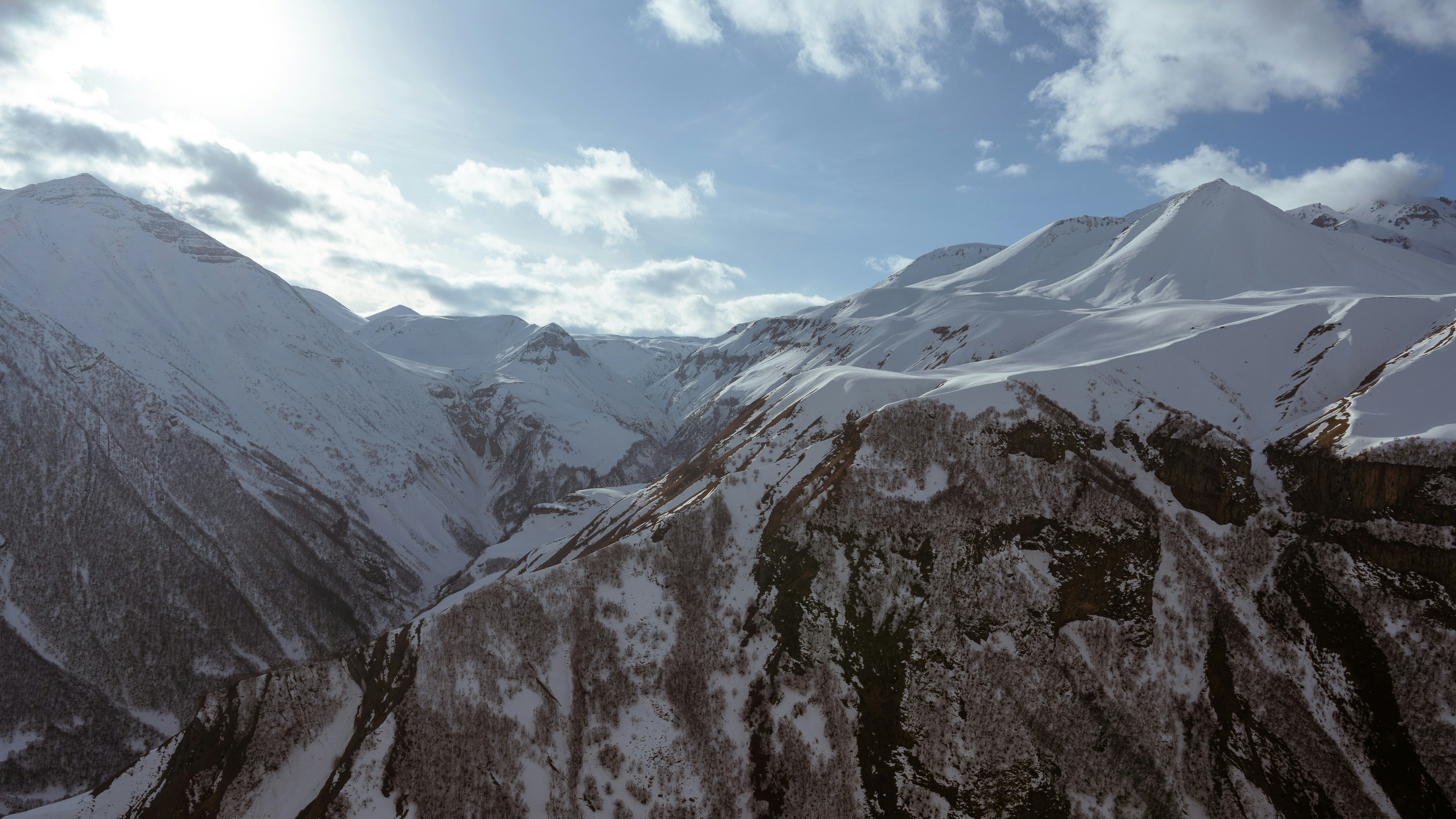 Snow-covered mountains under a partly cloudy sky with soft sunlight creating contrasting shadows.
