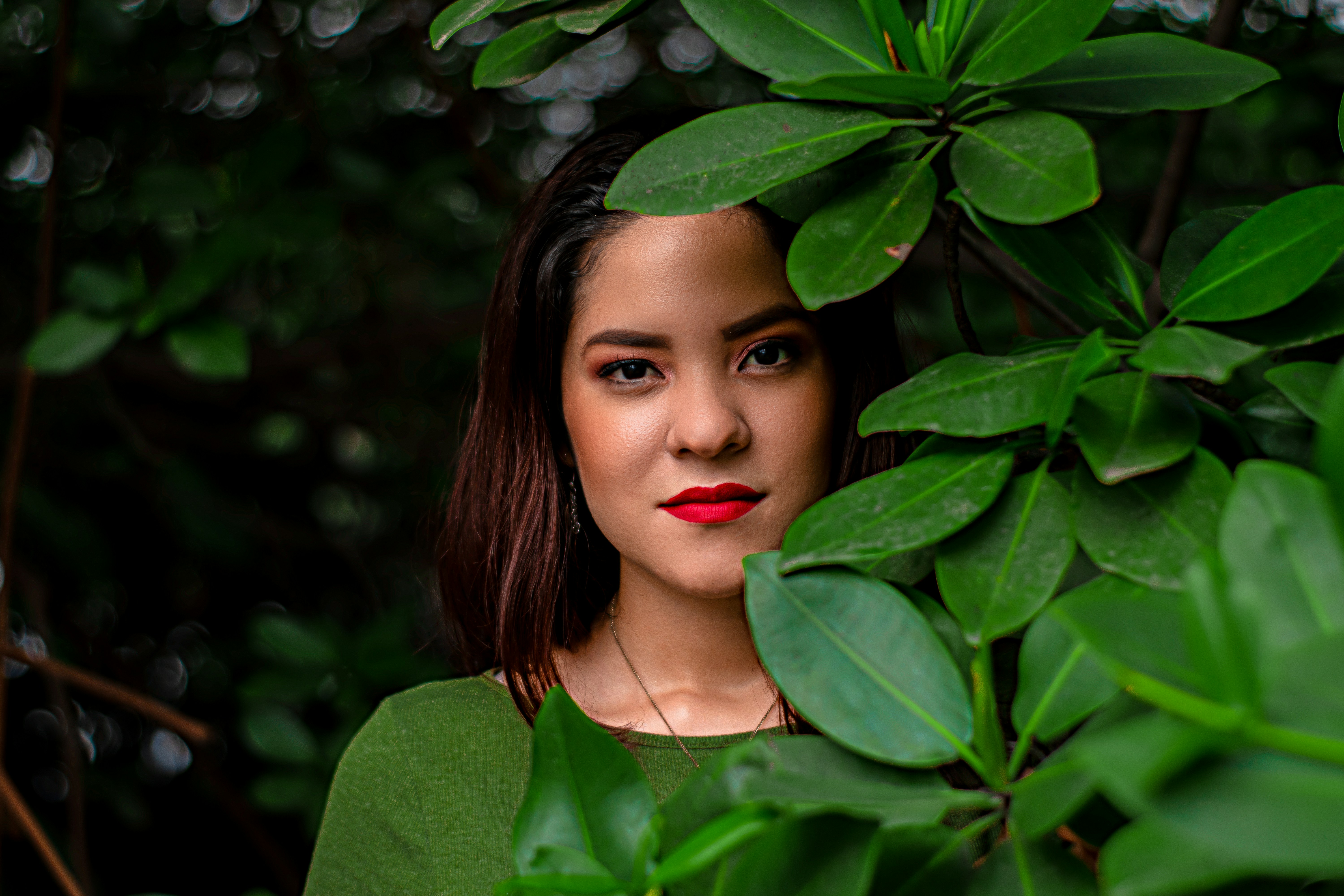Woman with red lipstick framed by vibrant green leaves, her expression serene amidst soft natural light.