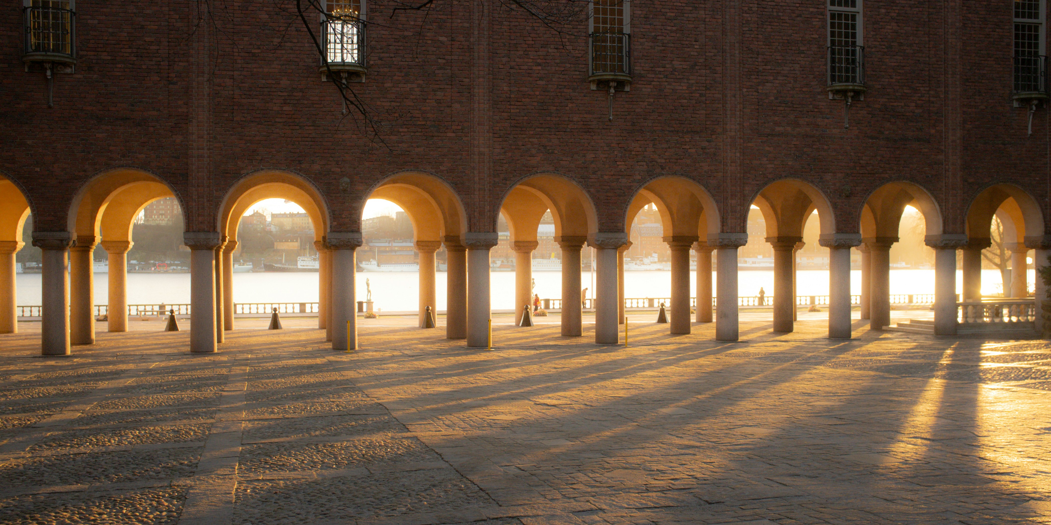 Sunlight streams through a row of arches, casting elongated shadows on a cobblestone courtyard.
