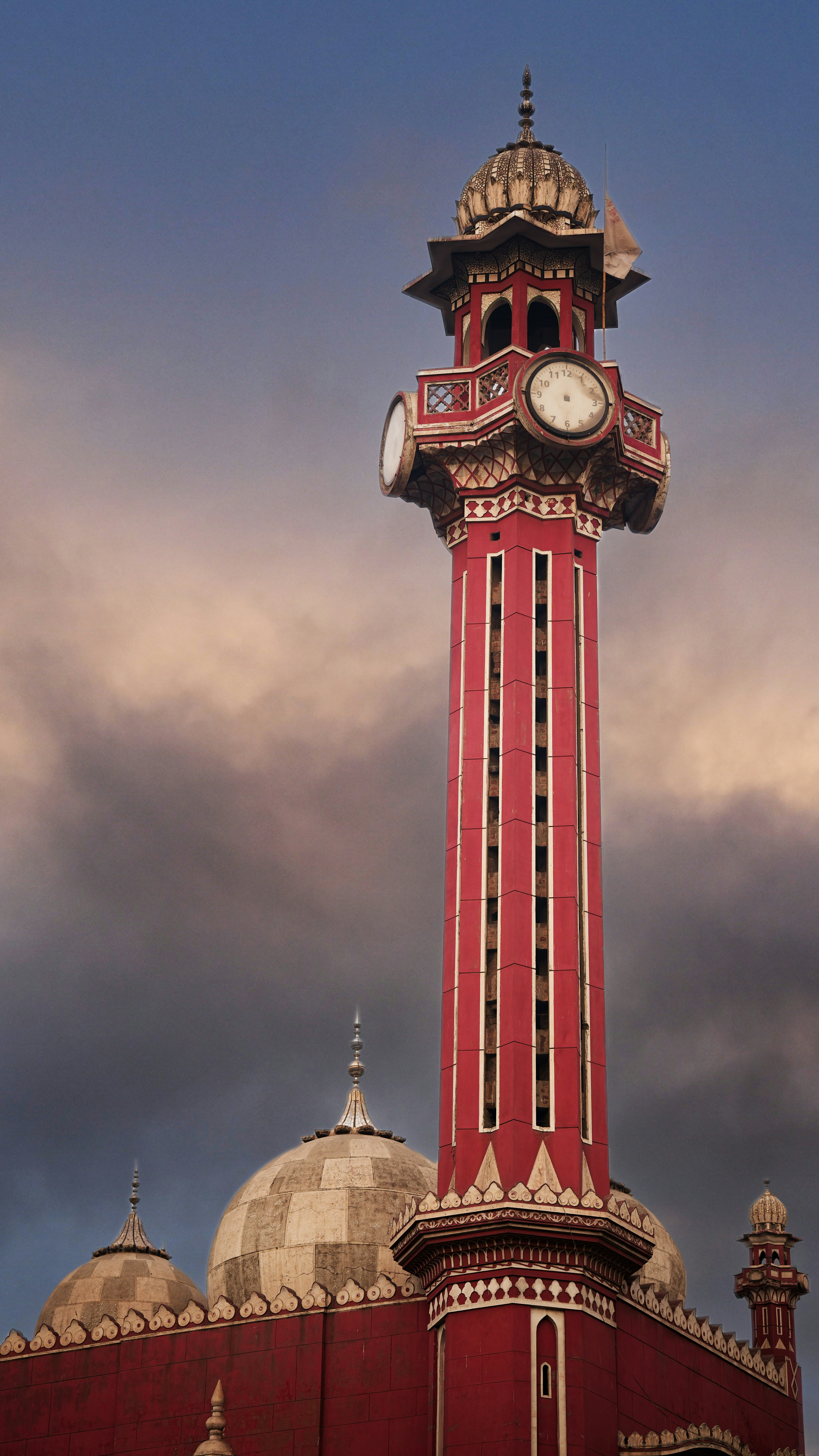 Red clock tower adorned with intricate designs rises against a moody sky, showcasing historical architecture. The tower's clock face stands prominently at the top.