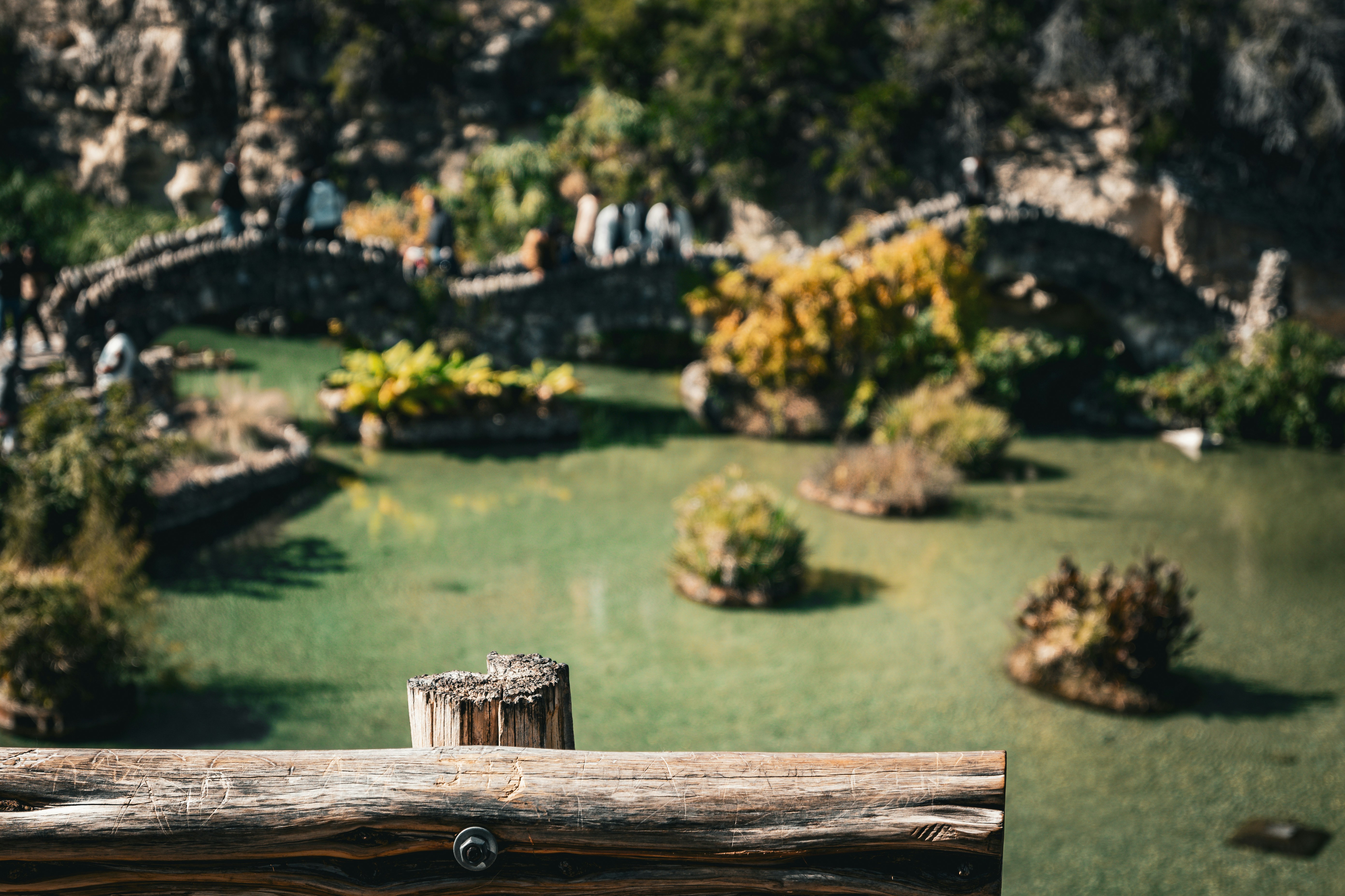 A rustic wooden railing in focus with a blurred stone bridge and lush greenery reflected in tranquil water.