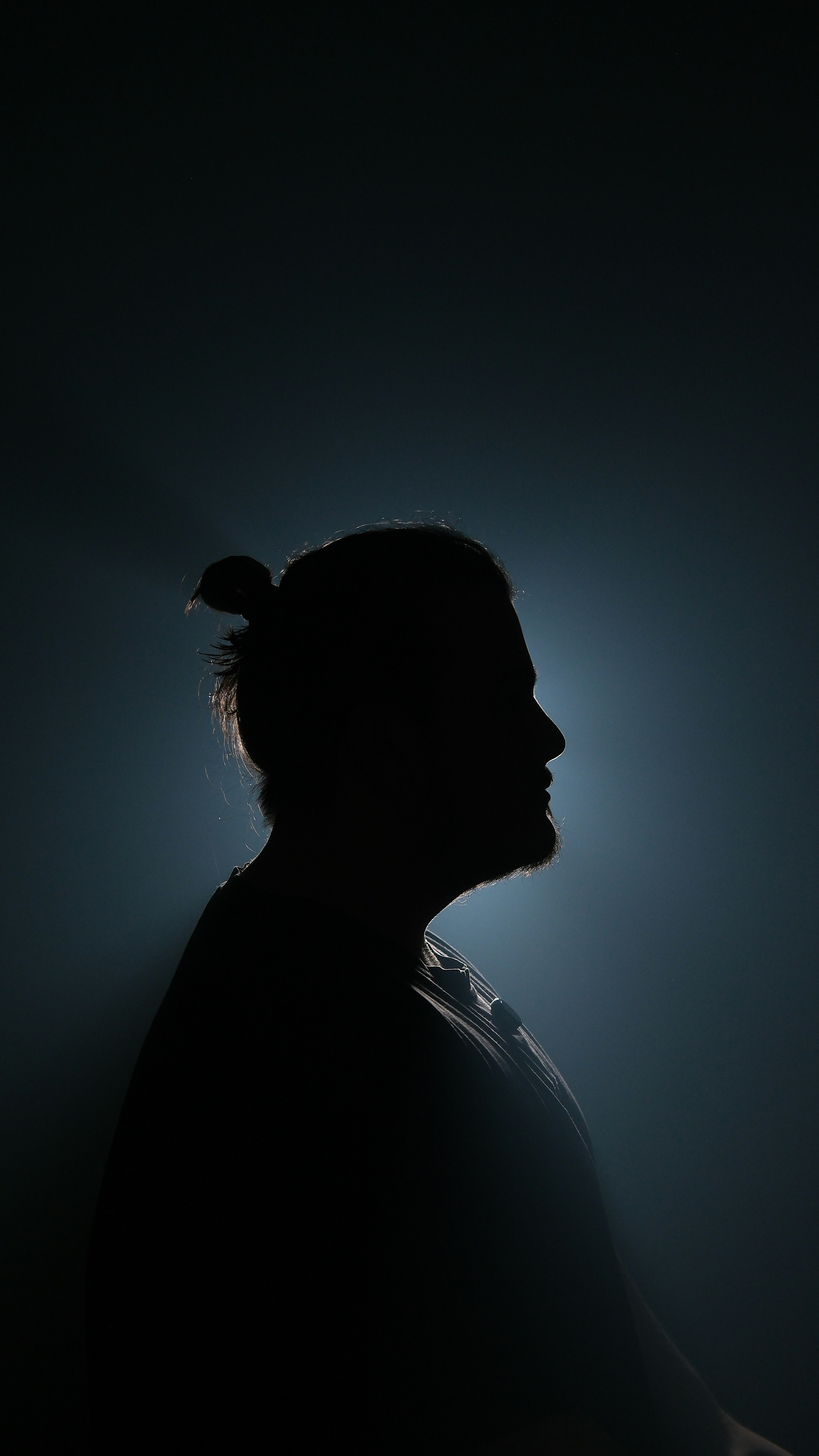 A dramatic silhouette of a man with a bun hairstyle, captured against a dark background with subtle light rays creating a halo effect around his head. The high-contrast lighting emphasizes the contours of his face and beard, adding depth and mystery to the composition. His relaxed posture and the minimalist setting evoke a sense of introspection and solitude. | A silhouette of a person in the dark