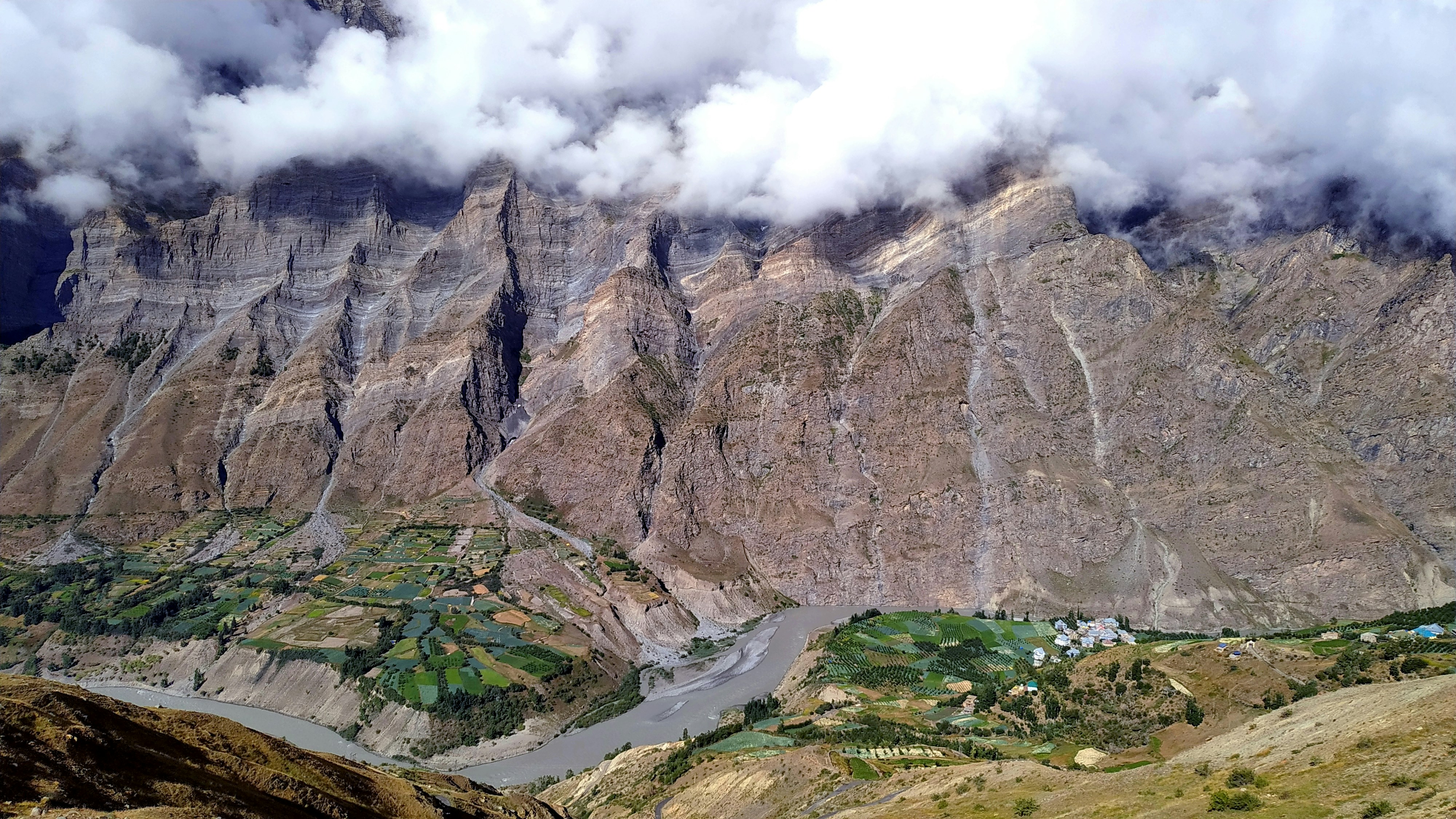 Towering barren mountains partially shrouded in clouds overlook lush fields and a winding river.