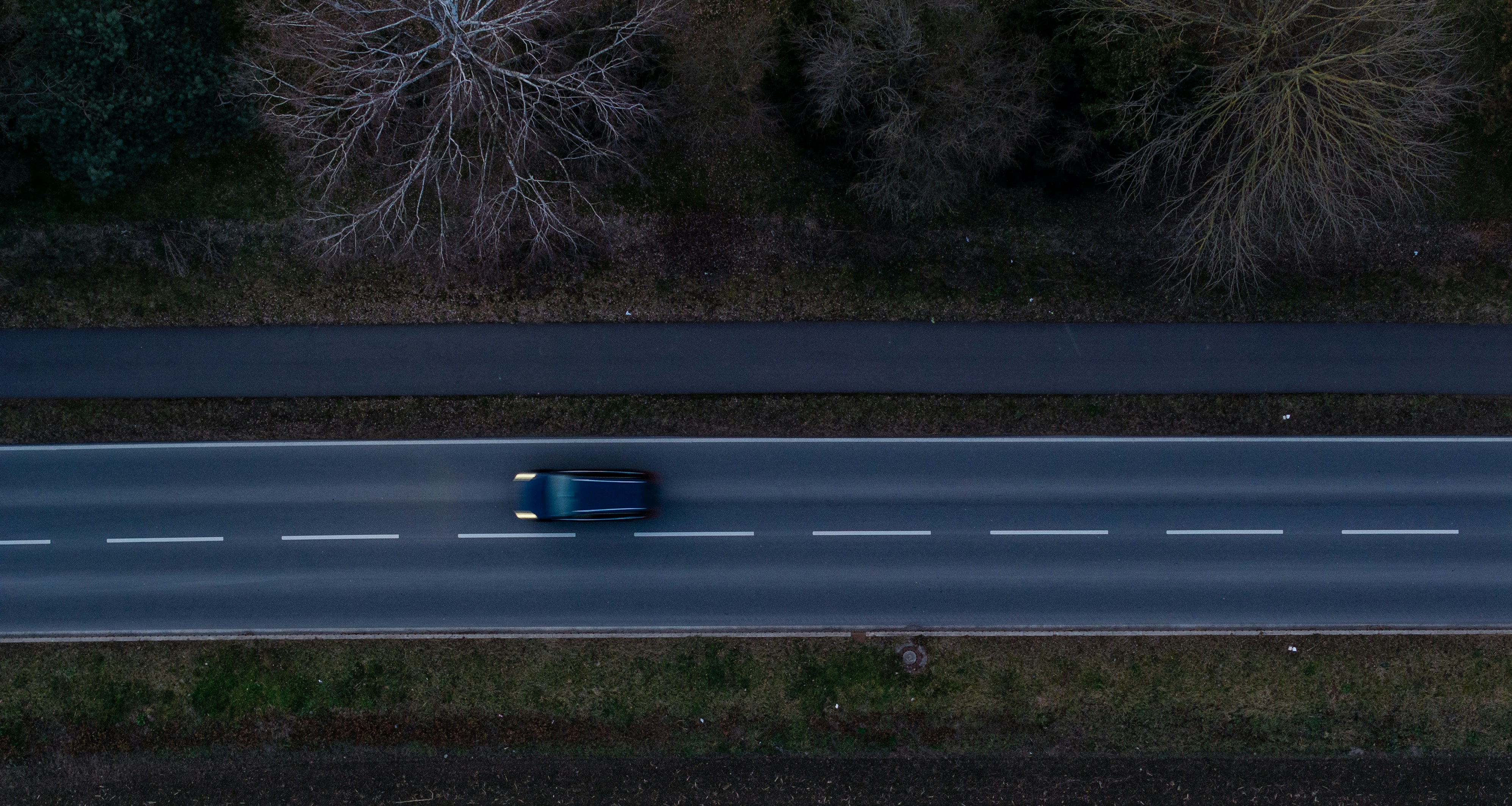 A lone car blurs along a straight road bordered by leafless trees and muted greenery.