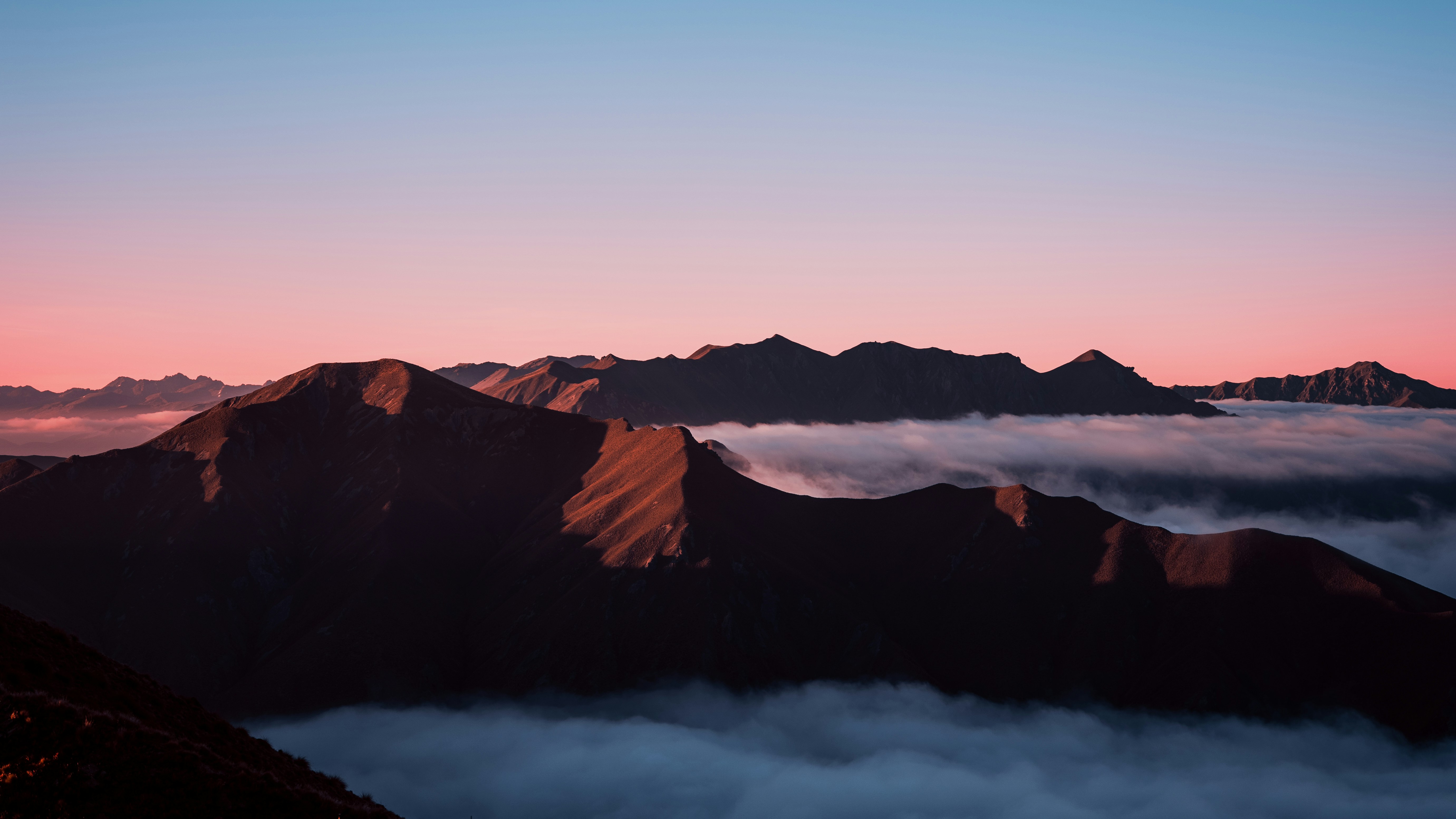 Mountain peaks silhouetted against a pastel dawn sky with clouds enveloping the lower slopes.