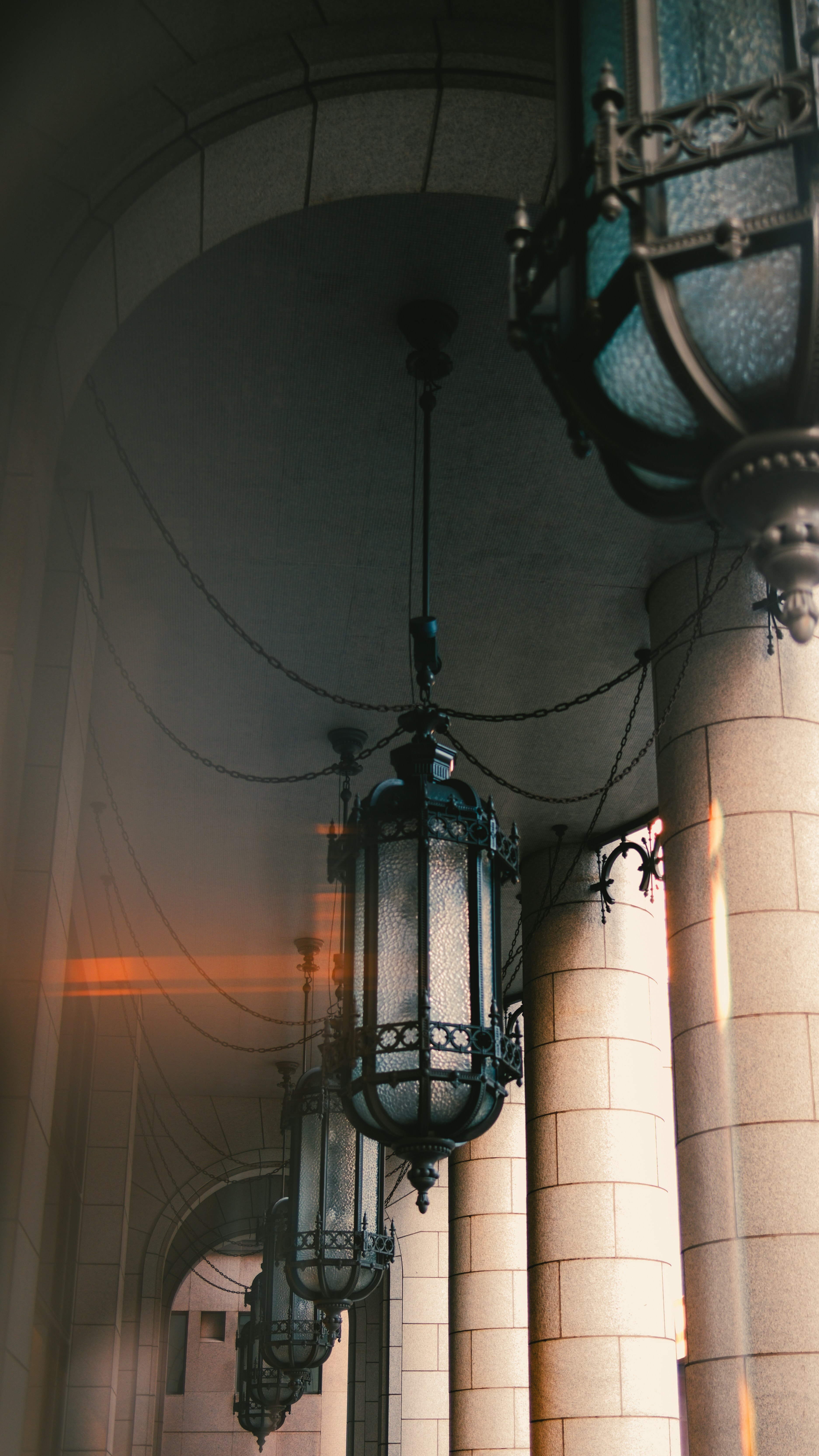 Row of ornate lanterns suspended along a stone arcade, softly lit with moody warm tones. The photograph emphasizes architectural details and the quiet, timeless atmosphere.