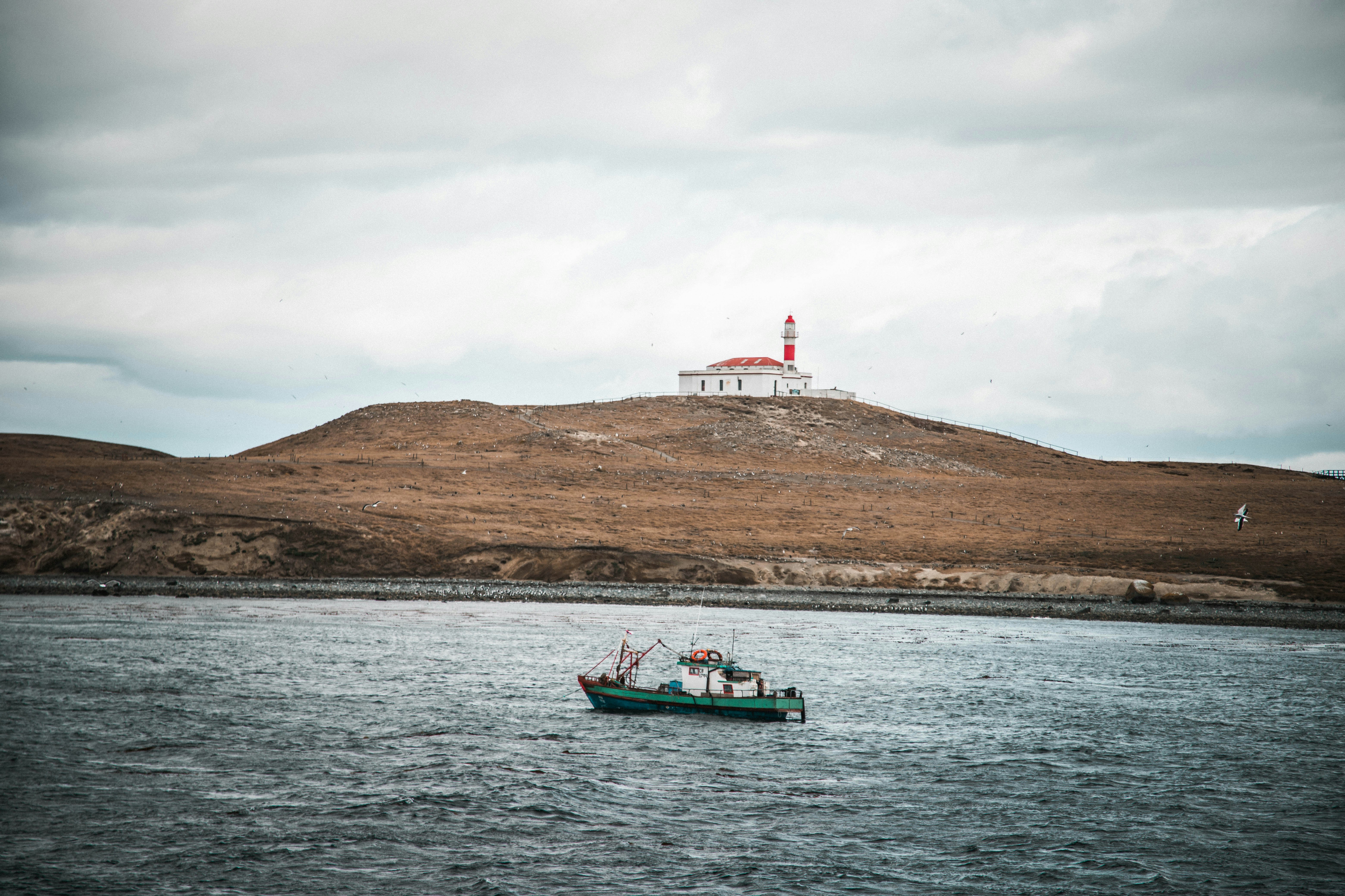 Lighthouse atop a barren hill under a cloudy sky, with a lone fishing boat on calm sea waters.