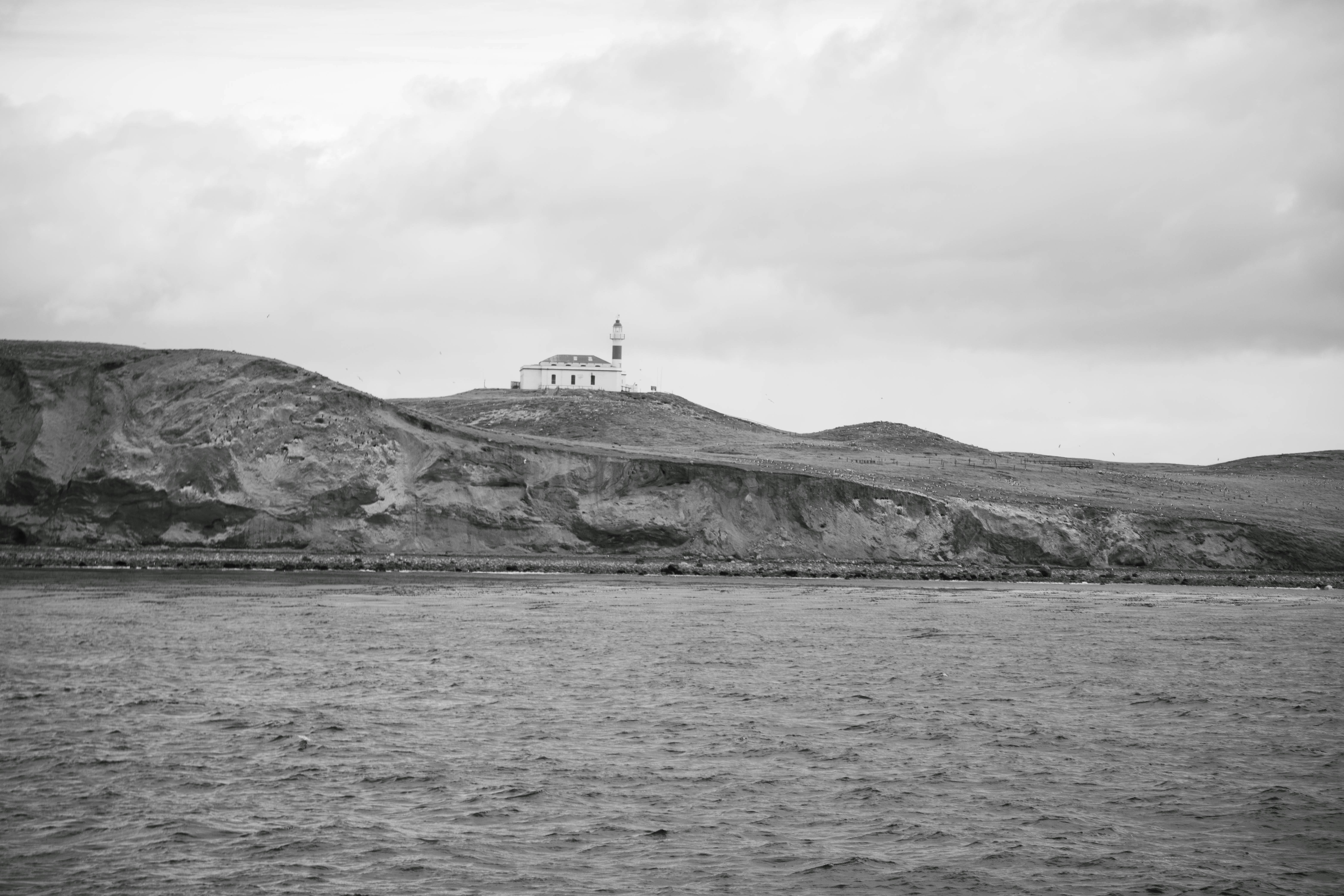 Monochrome scene of a lighthouse atop a cliff, overlooking a choppy sea under a cloudy sky.