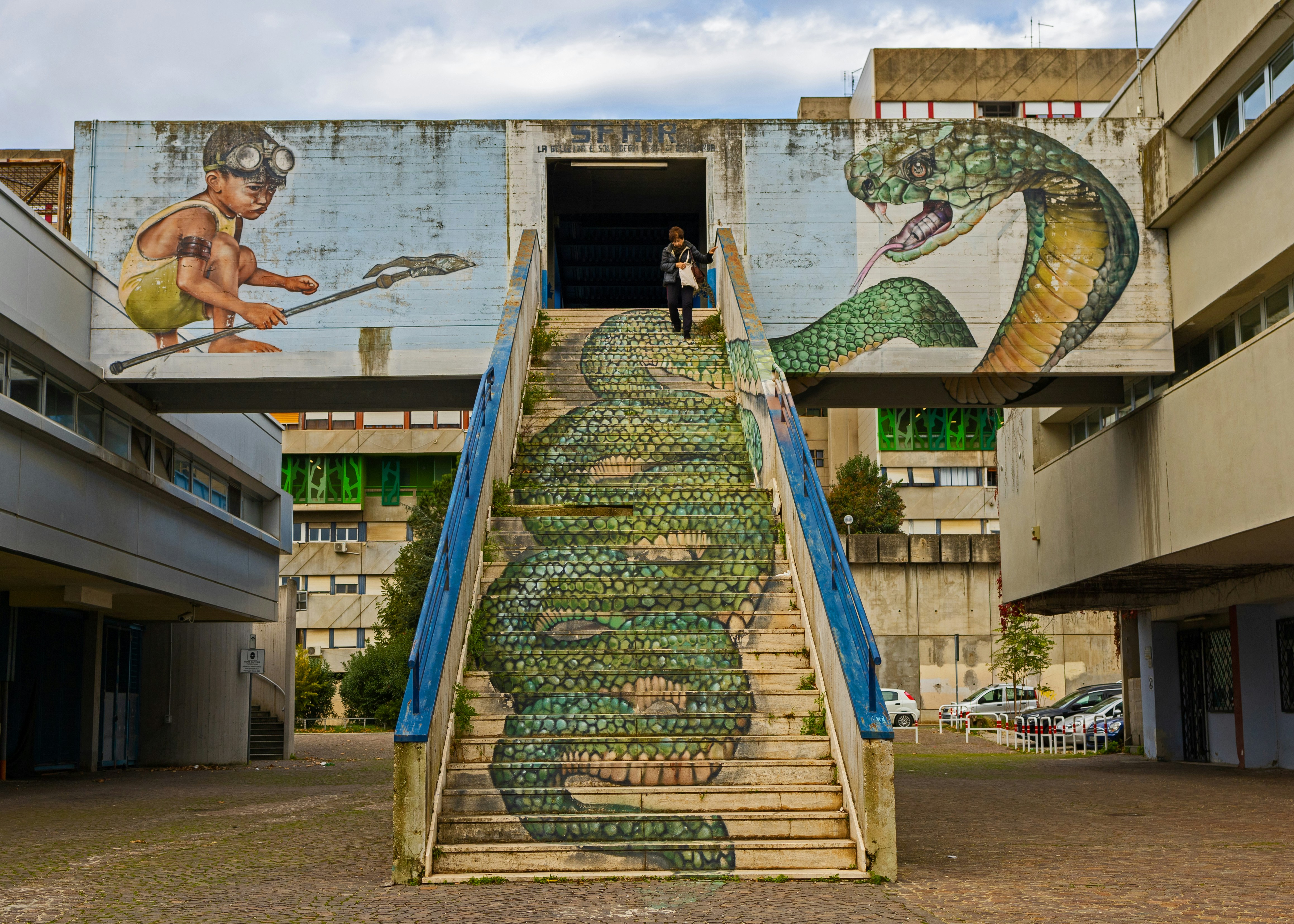 Vibrant mural of a serpent on a staircase with a child and snake artwork on the wall, set against weathered buildings.