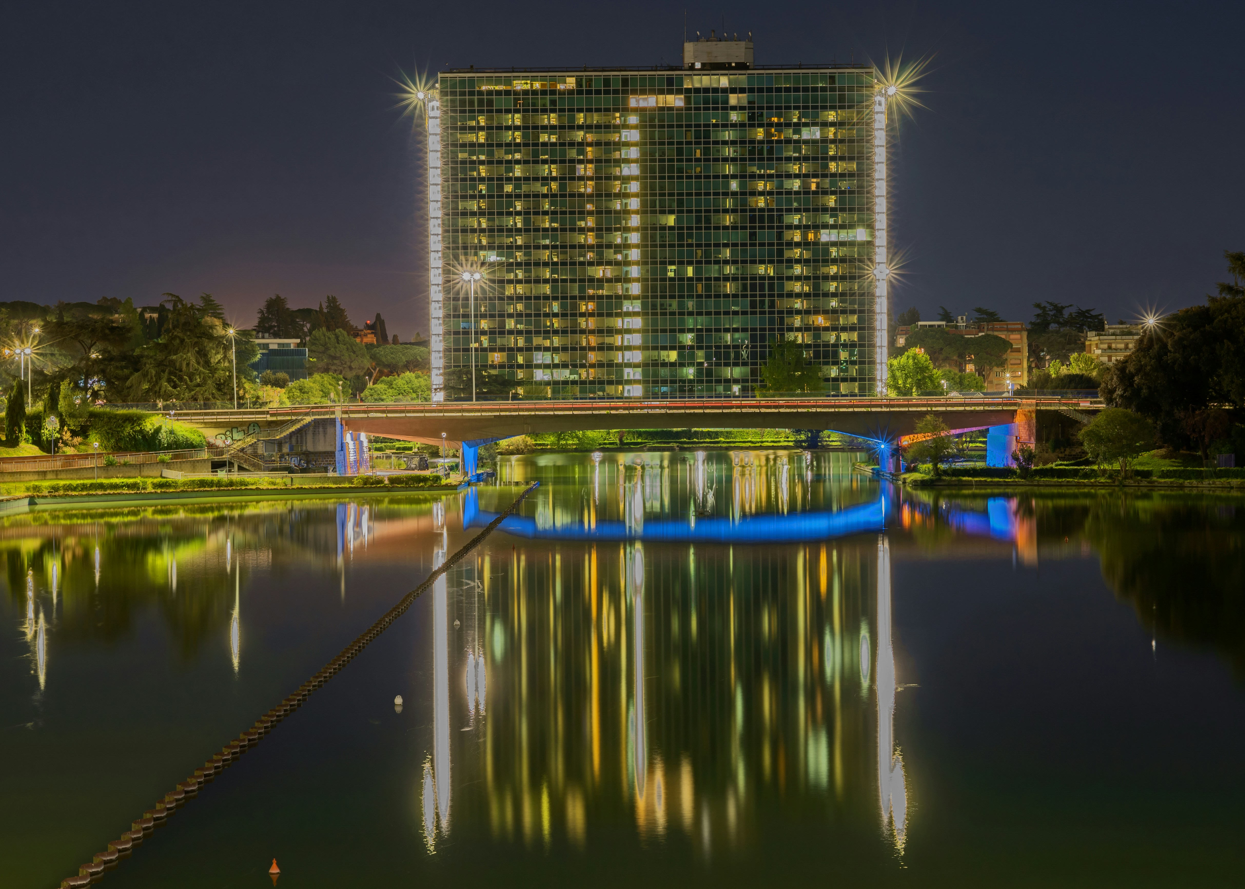 Illuminated skyscraper reflecting on a calm river under a starry night sky with surrounding greenery.