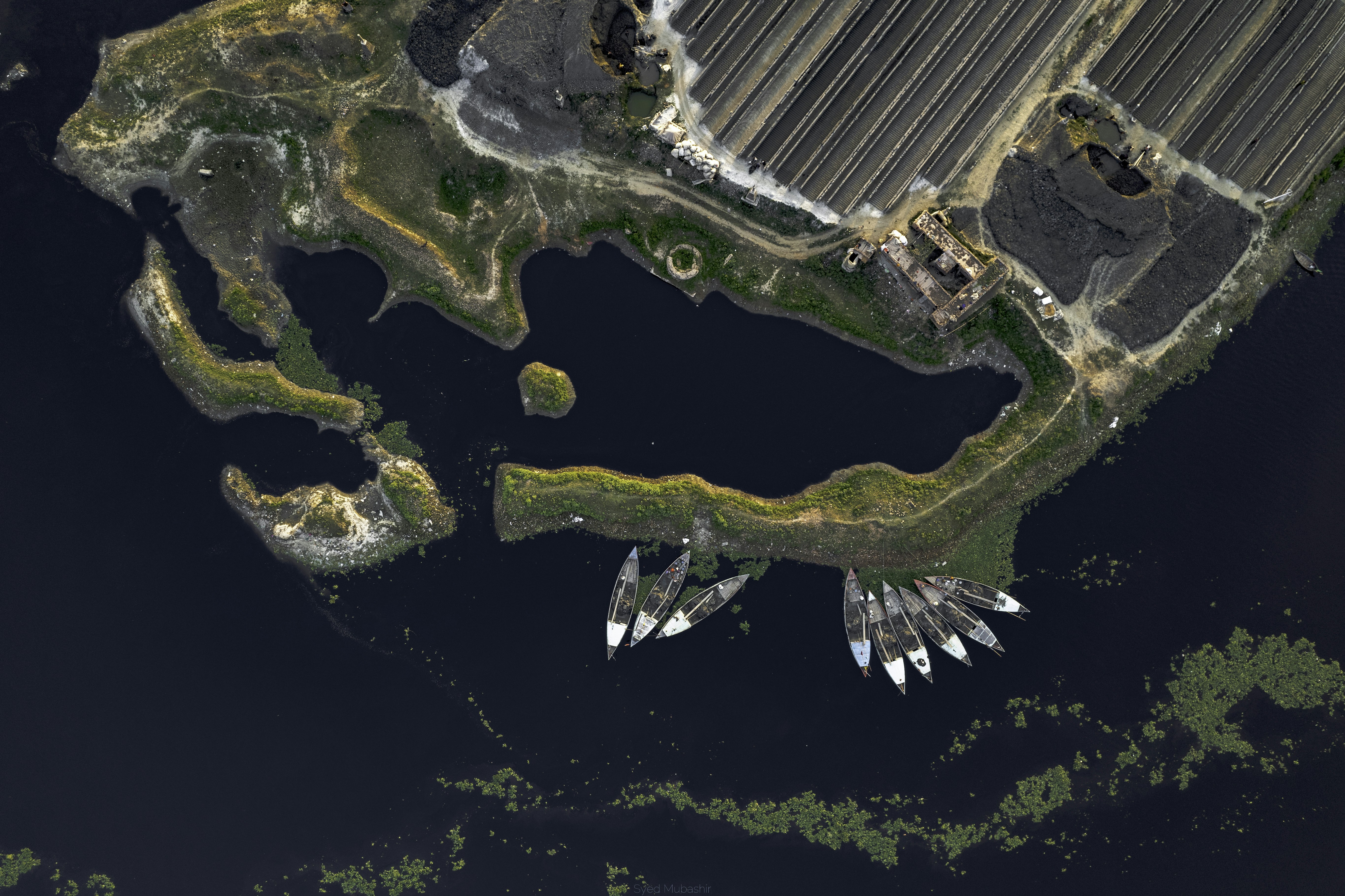 Aerial view of boats aligned along a lush green shoreline, bordered by deep blue water and earthy landscapes.
