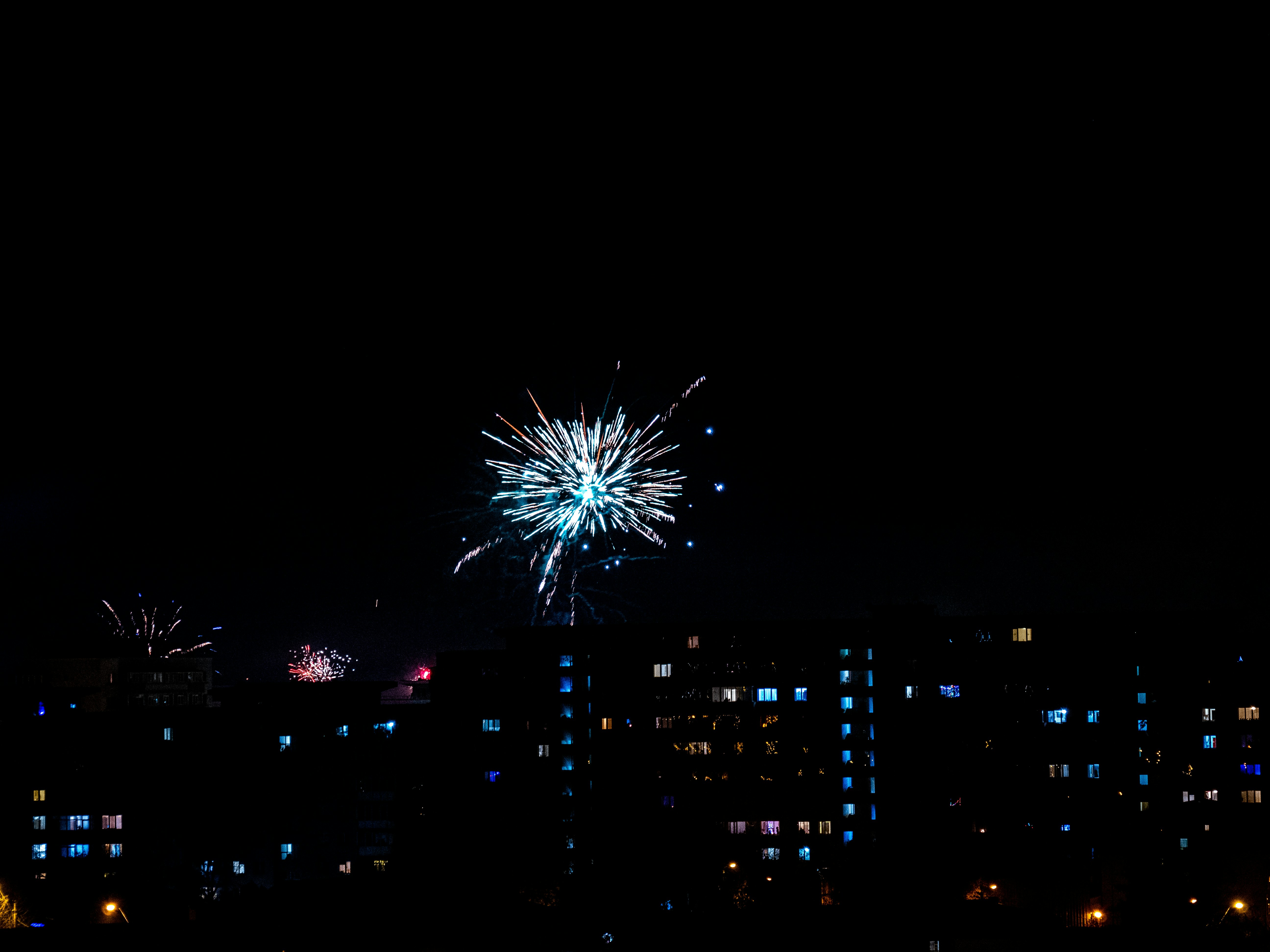 Fireworks burst in vivid blue and white against a dark city skyline, with illuminated windows adding depth.
