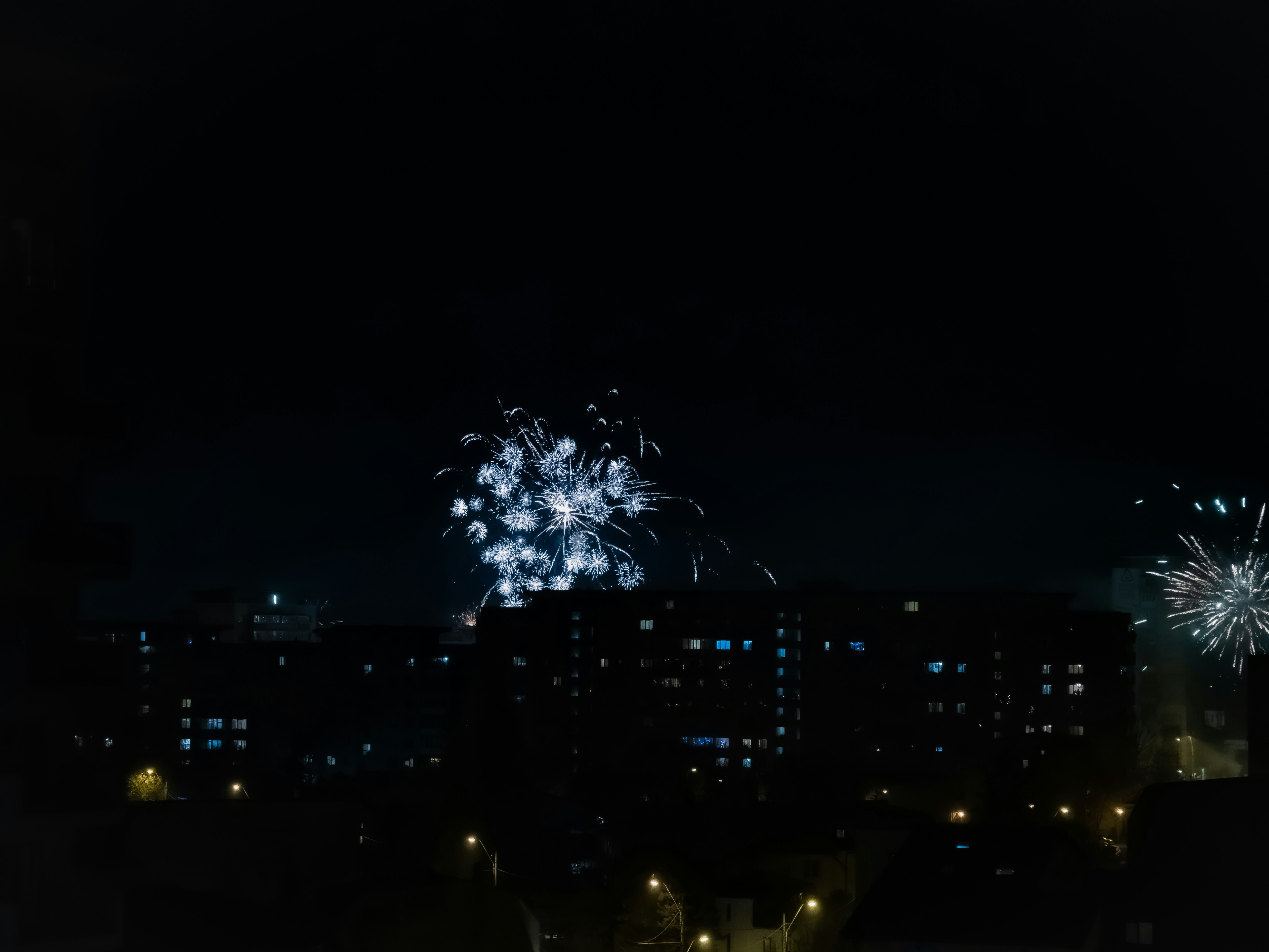 White and blue fireworks illuminate the night sky above dark city buildings, with streetlights casting a warm glow.
