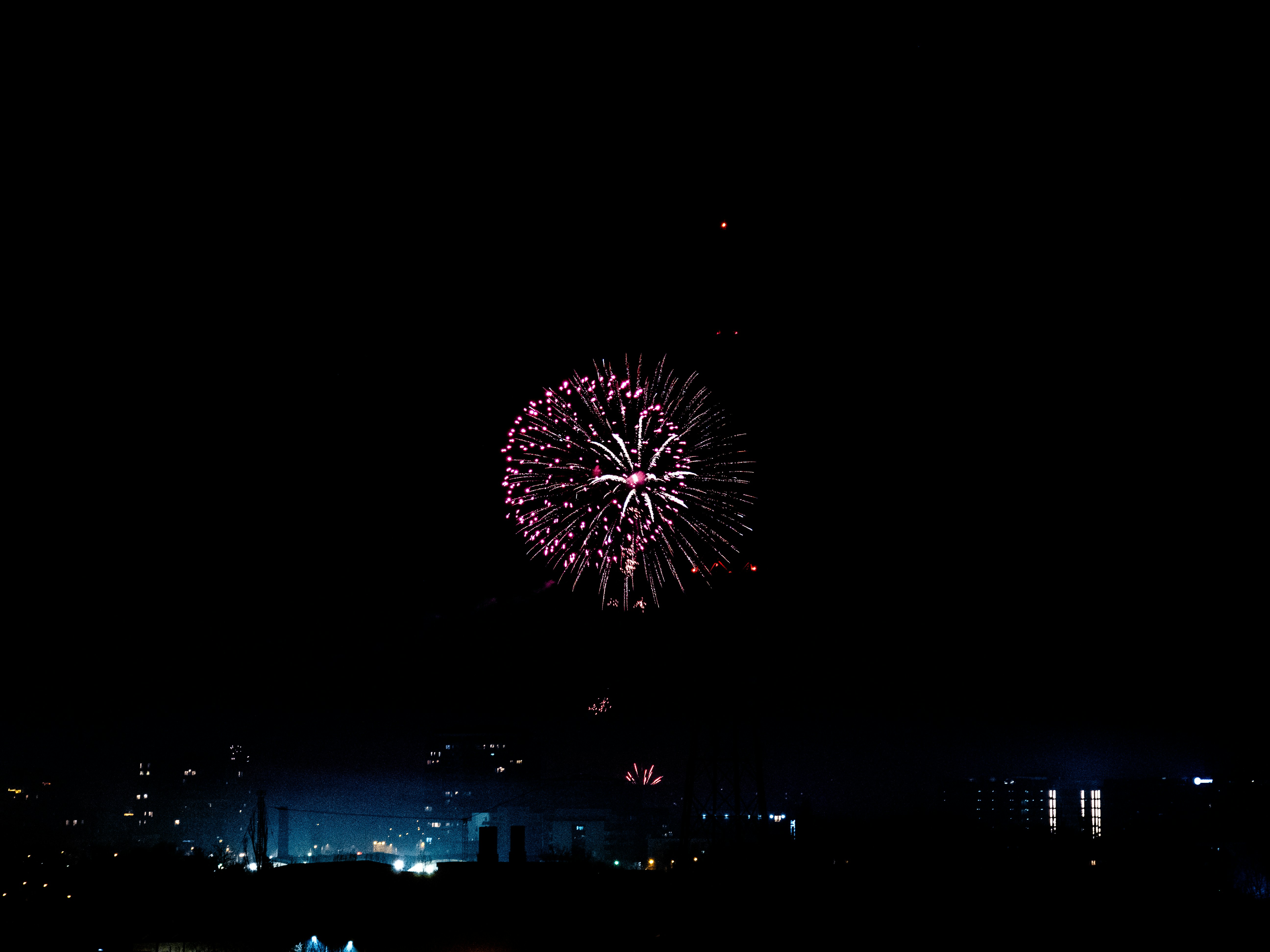 Fireworks burst with pink and red hues against a dark night sky above a silhouetted cityscape. Subtle city lights below add depth and atmosphere.