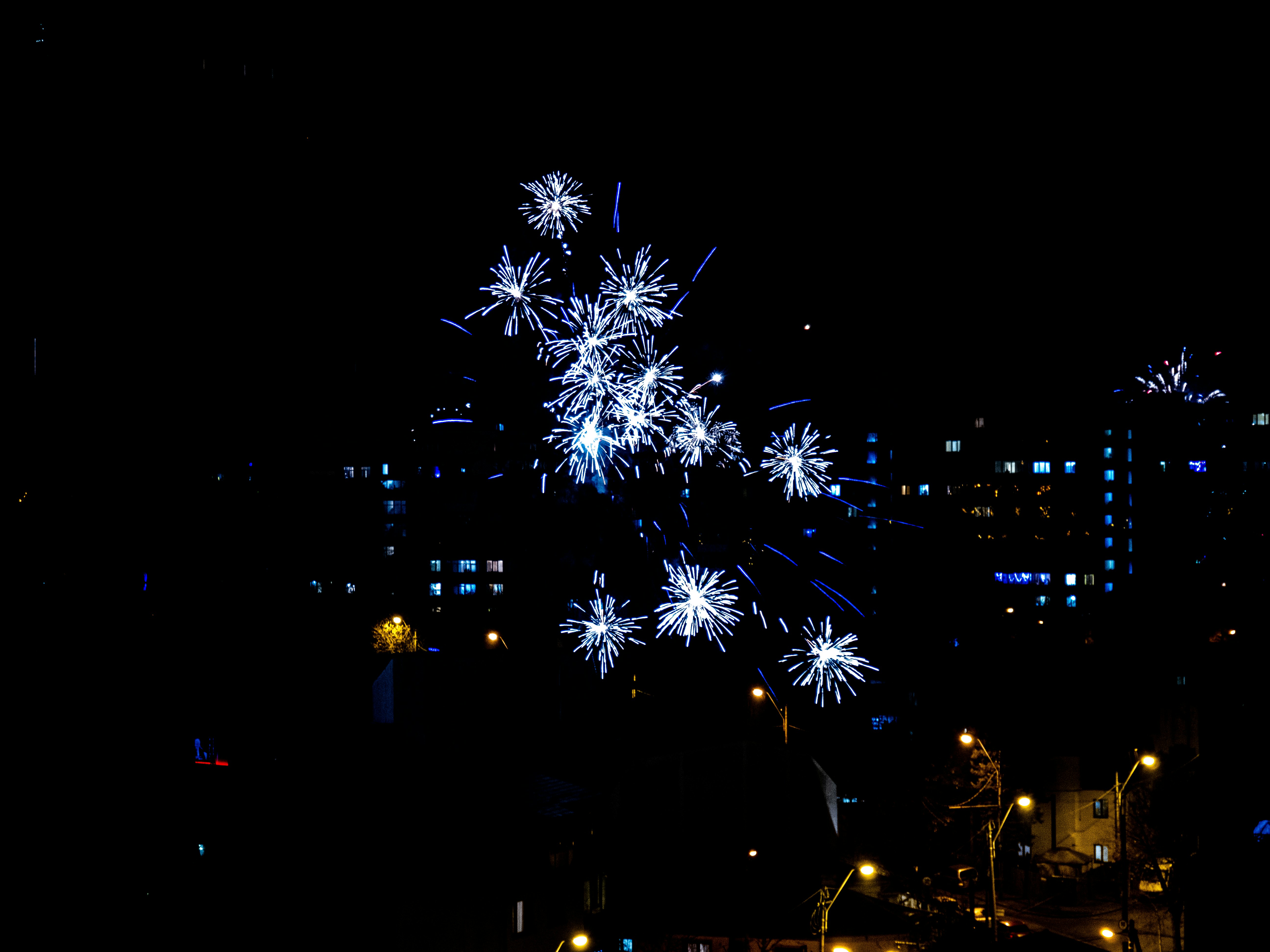 White and blue fireworks burst over a dark cityscape, illuminating urban structures below.