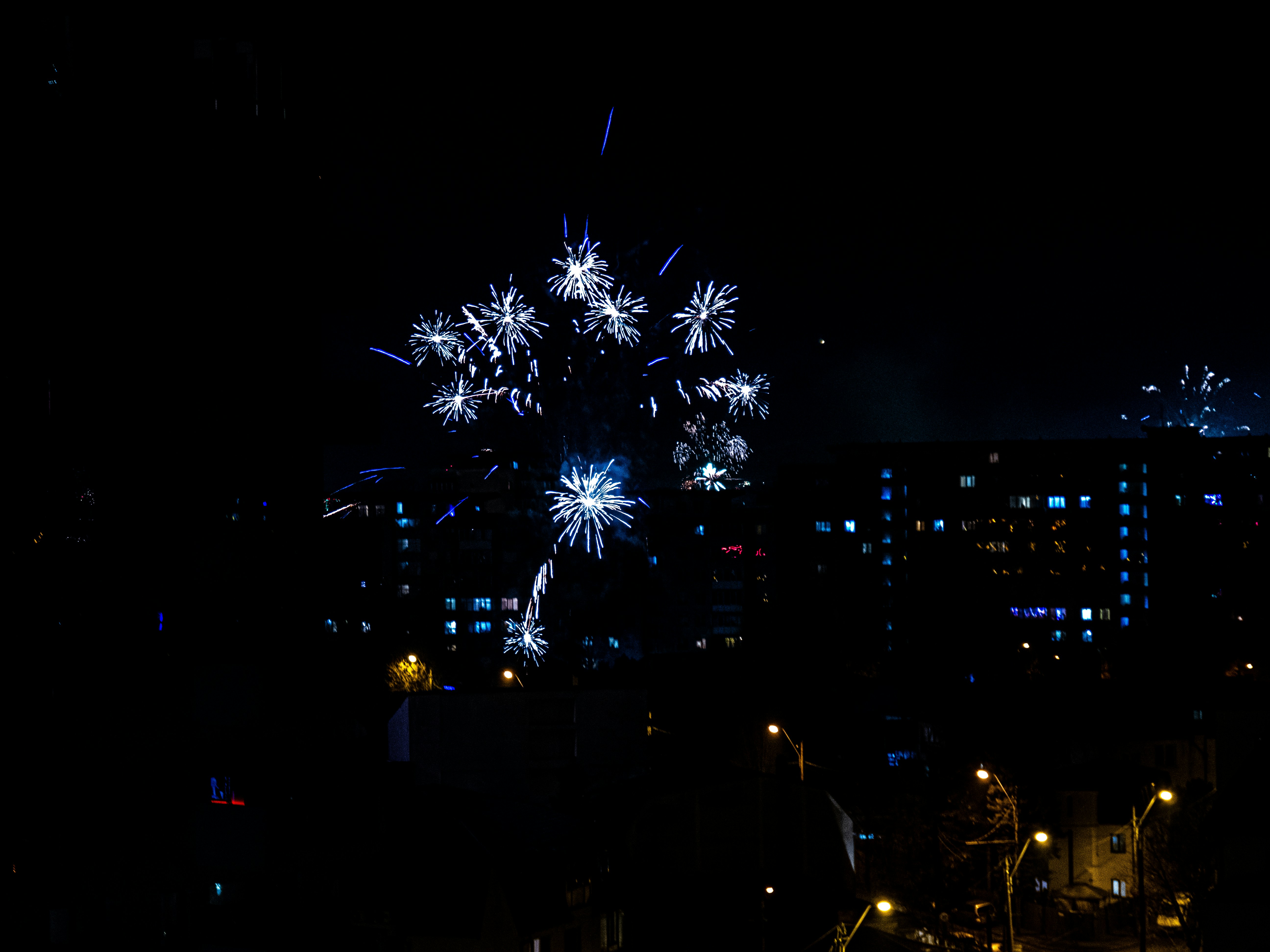 White and blue fireworks erupt over a dark cityscape, with streetlights casting a warm glow below.