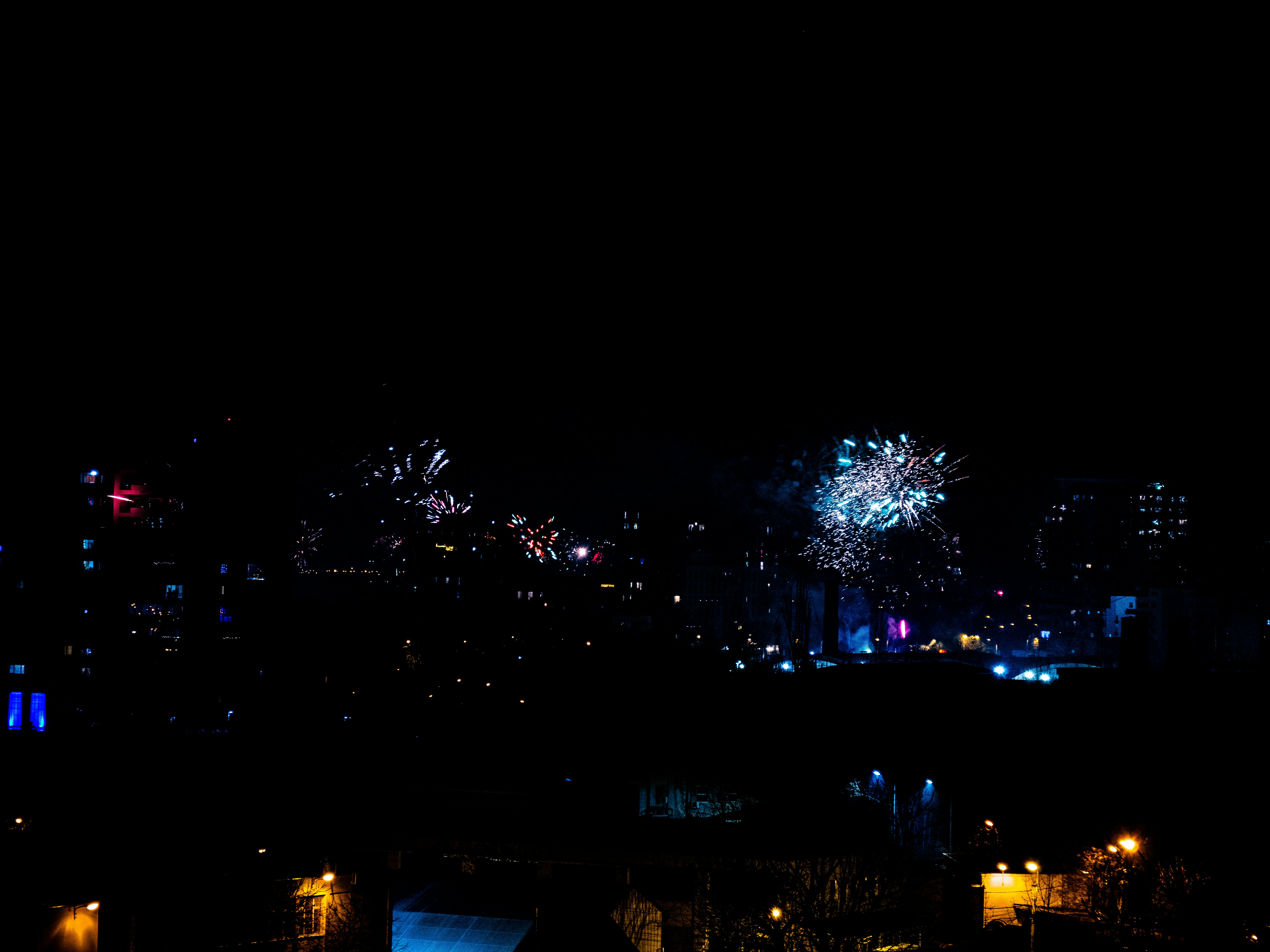 Fireworks burst in blue and white over a dark cityscape, with urban lights twinkling below.