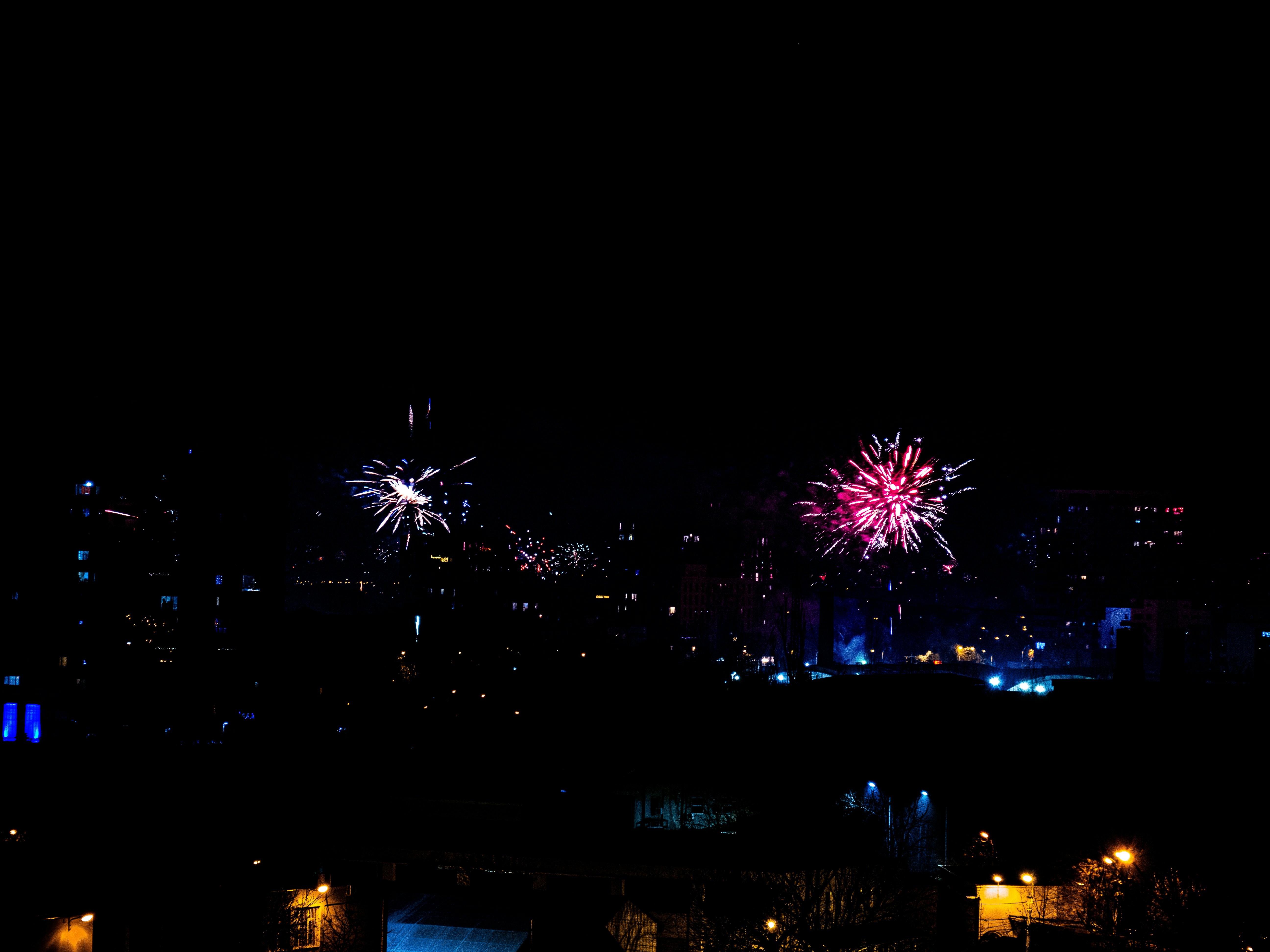 Fireworks burst in red and white above a silhouetted cityscape against a deep indigo sky.
