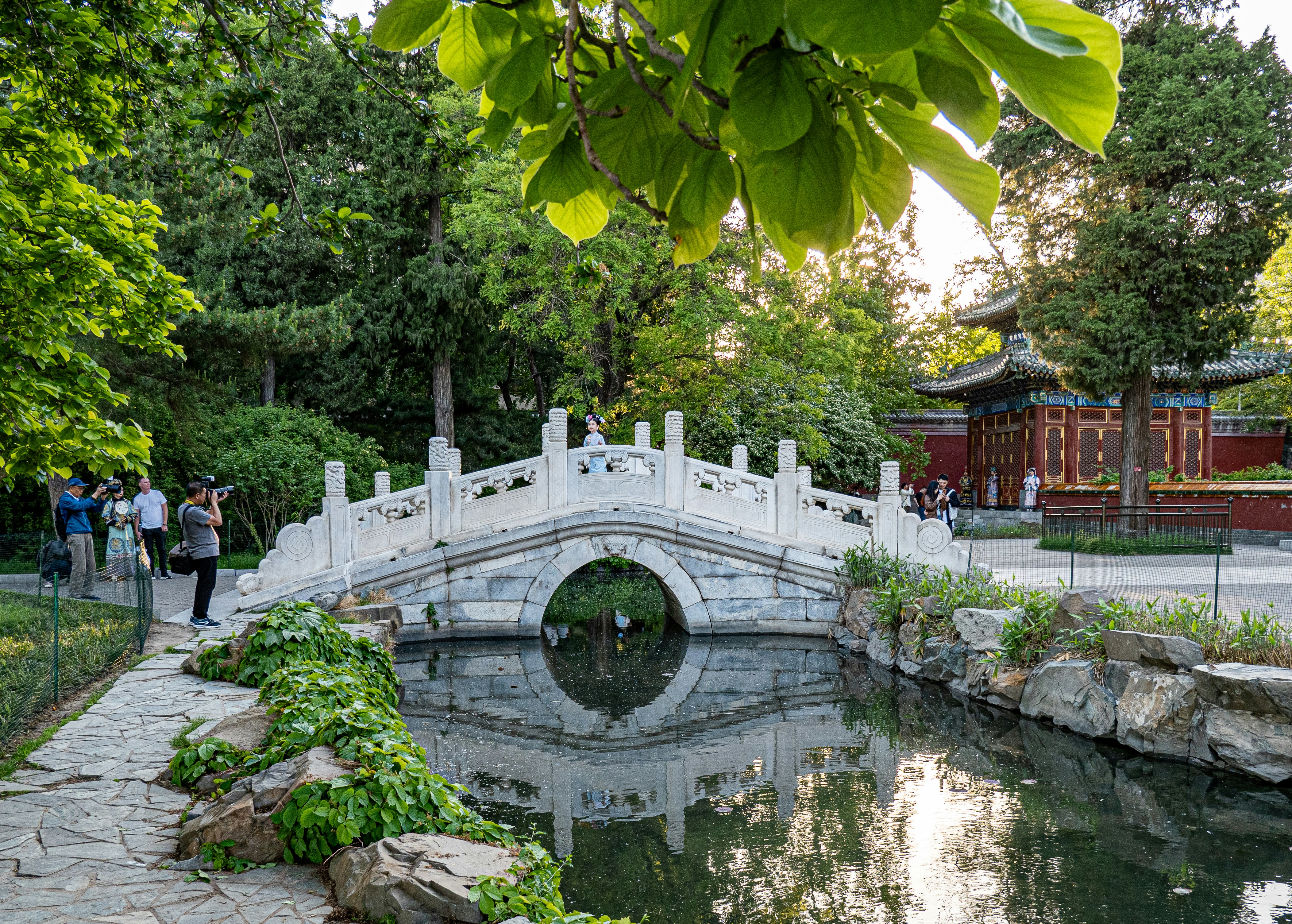 Stone bridge arches over a reflective pond with lush greenery and traditional architecture in the background.
